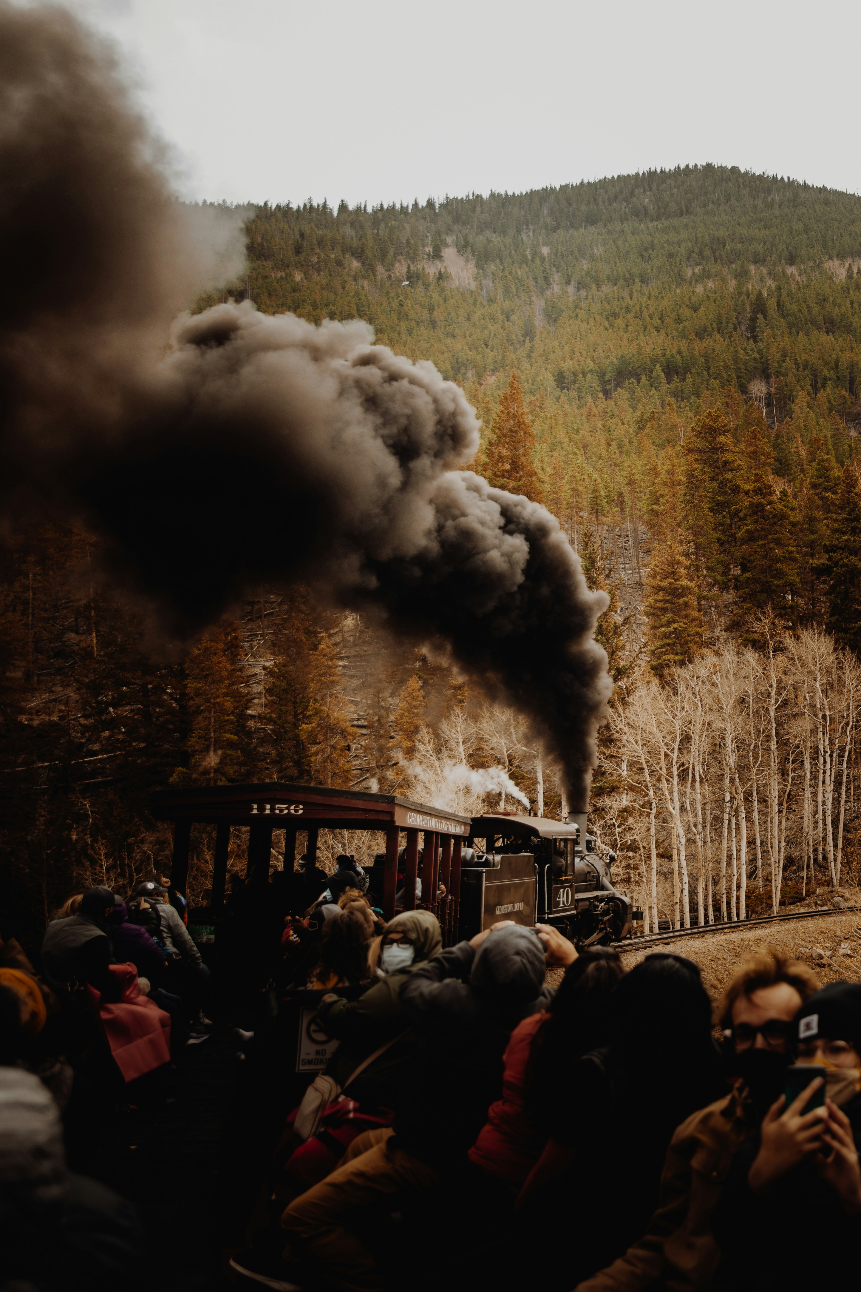 Steam locomotive billowing smoke as it travels through forested hills, surrounded by a crowd of onlookers.