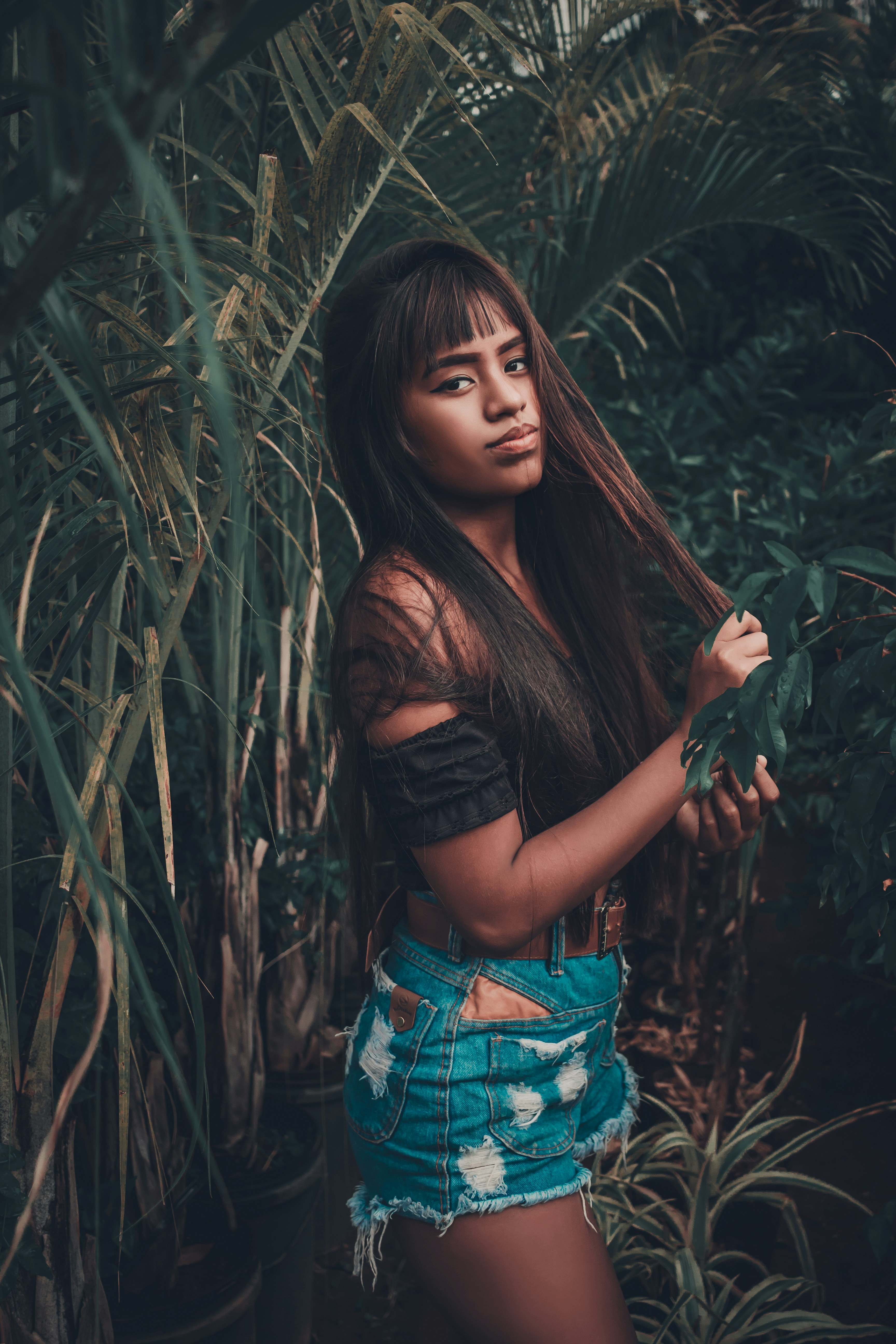 Woman in casual attire standing amidst lush tropical foliage with a contemplative expression.