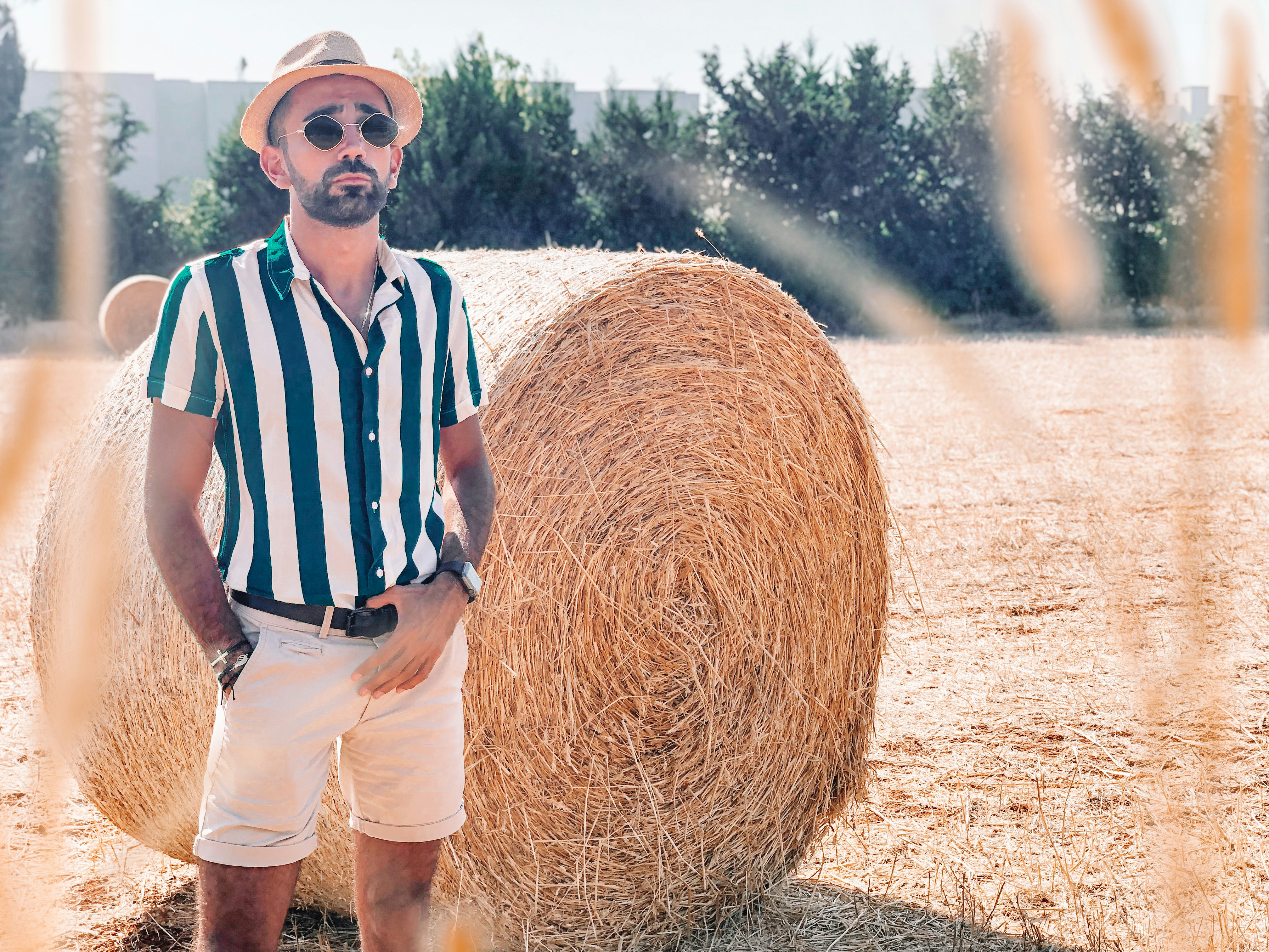 A man in a striped shirt and hat stands confidently next to a large hay bale in a sunlit field. The scene captures a relaxed summer vibe.