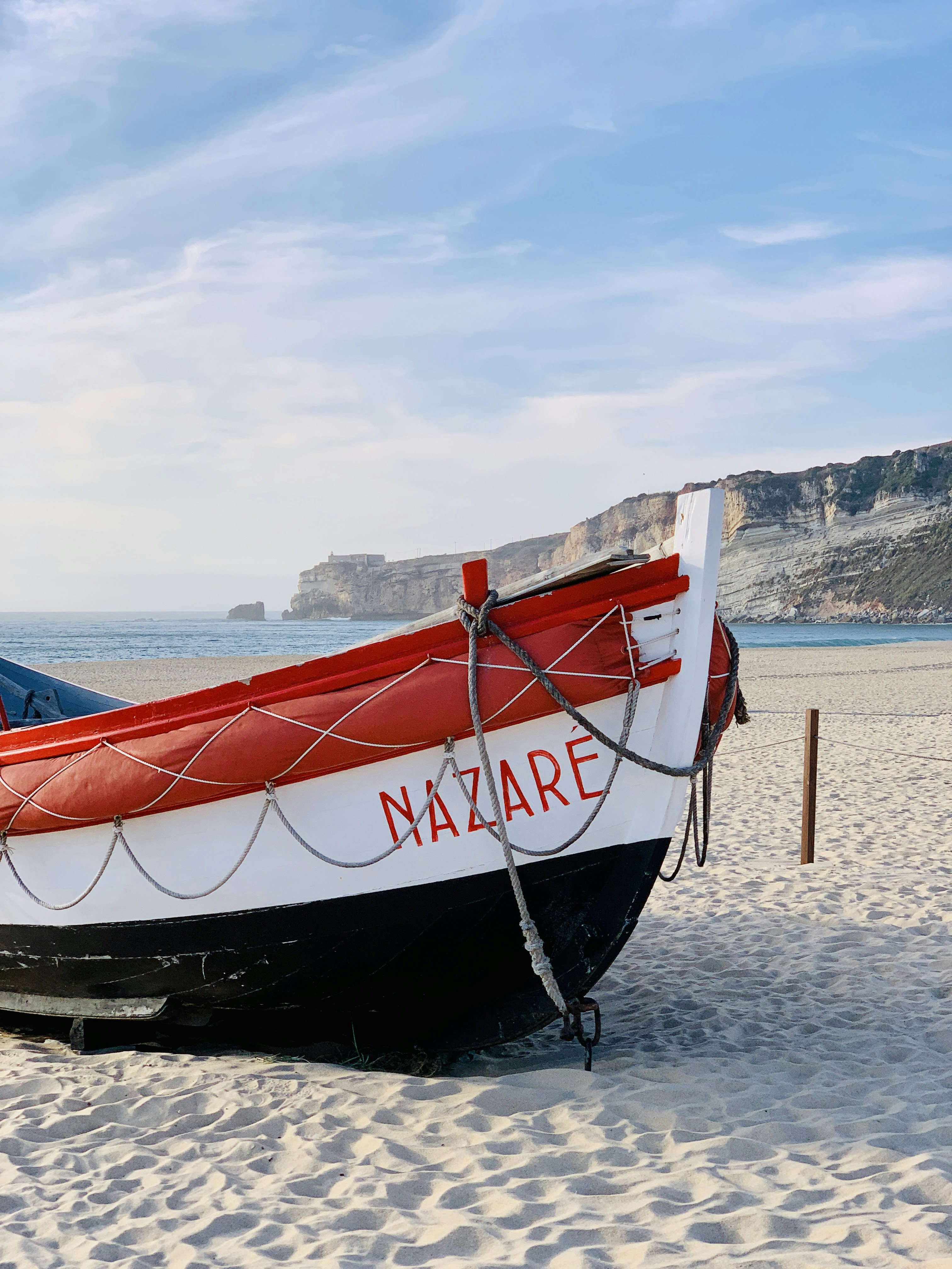 Vibrant fishing boat named 'NAZARE' resting on sandy beach with cliffs in the background. The scene reflects the coastal charm and maritime culture of the region.