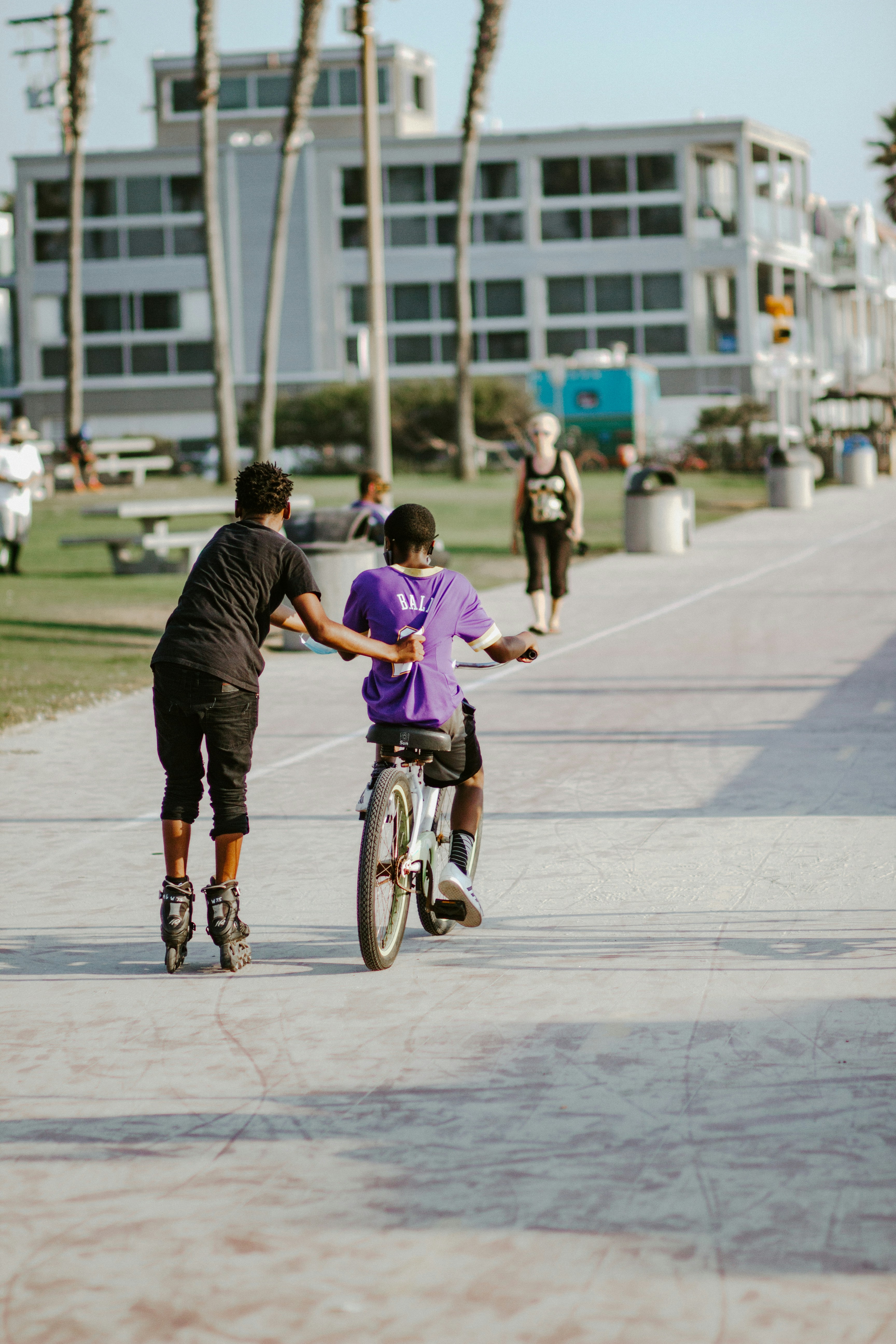 Two friends wheel a bike along a sunlit boardwalk while a pedestrian watches