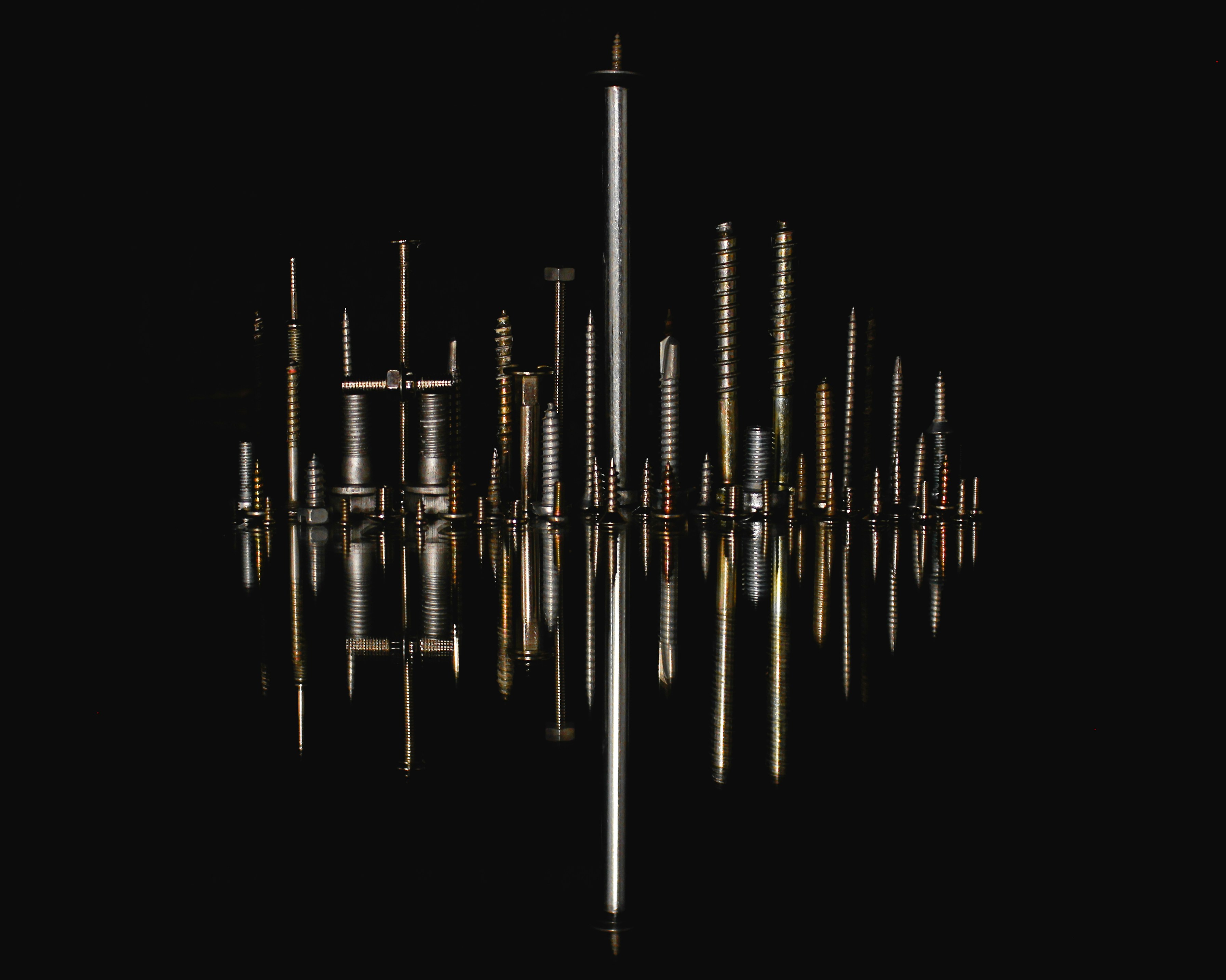 A collection of screws and bolts arranged in a striking display, reflecting on a dark surface. The interplay of light and shadow highlights the intricate details of each piece.