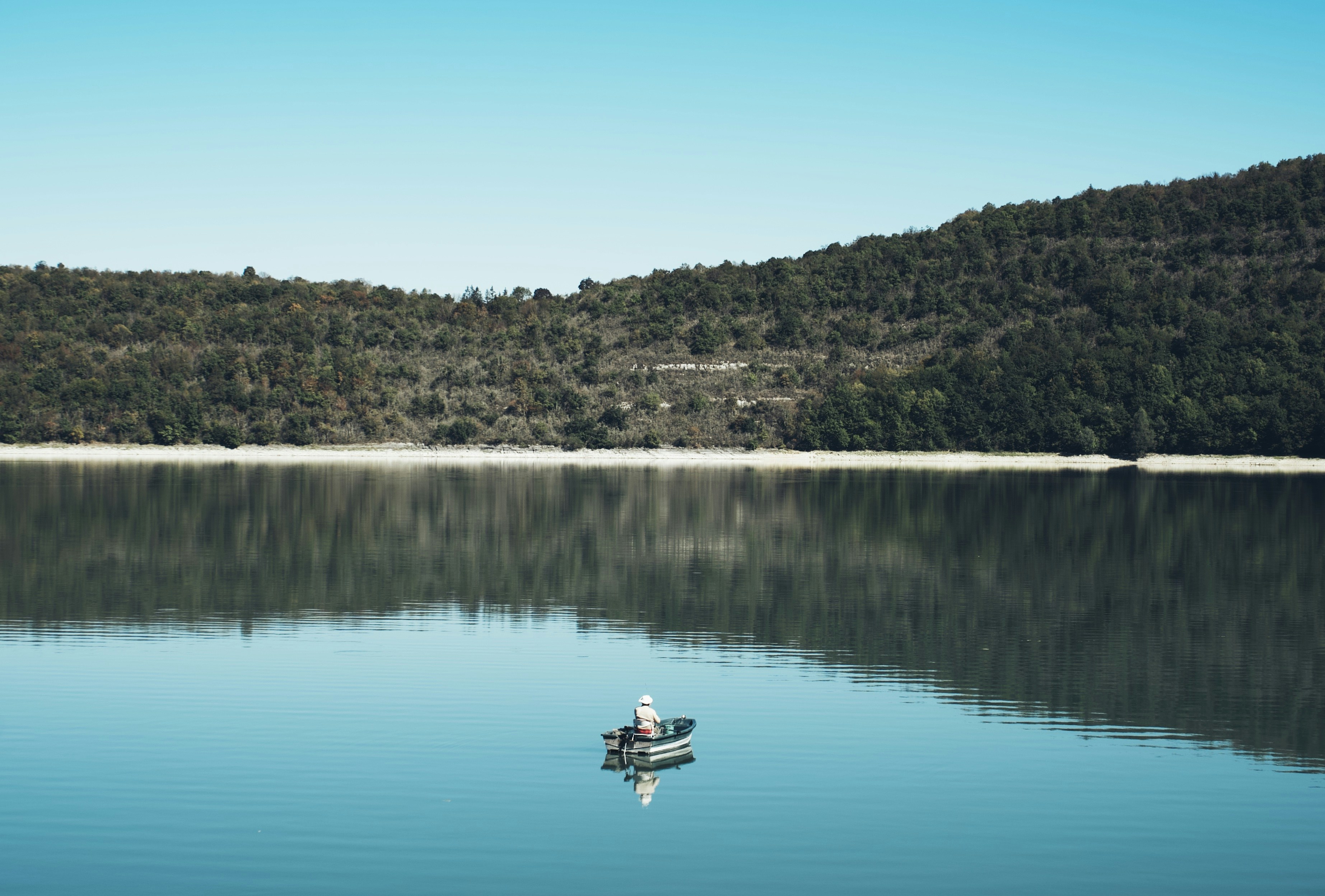 A lone fisherman in a small boat reflects on still waters, surrounded by lush green hills under a clear blue sky.