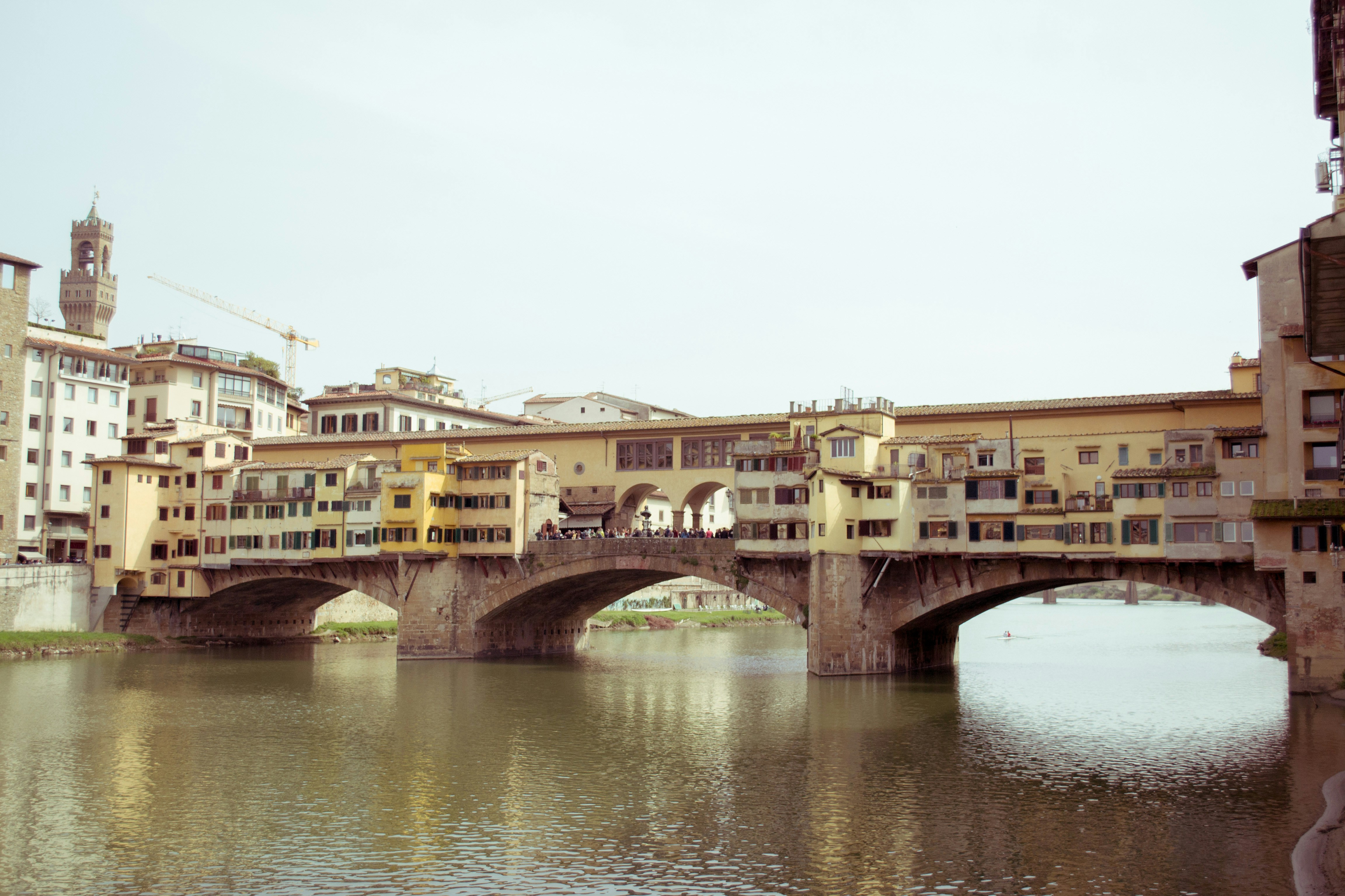 Historic bridge adorned with vibrant buildings spans a tranquil river, reflecting the charm of the surrounding architecture.