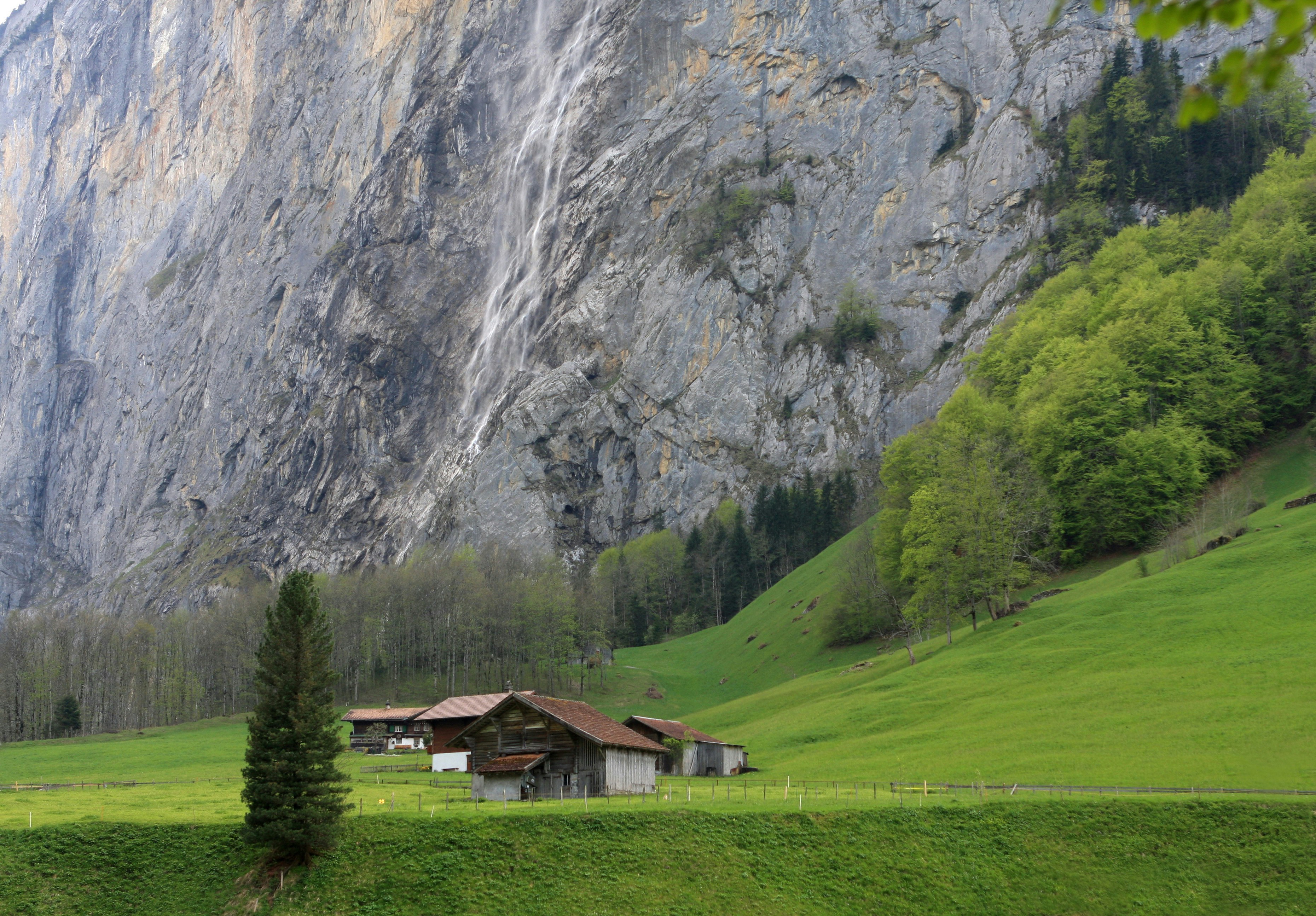 Charming wooden chalets nestled in a lush green valley beneath a towering cliff with a cascading waterfall.