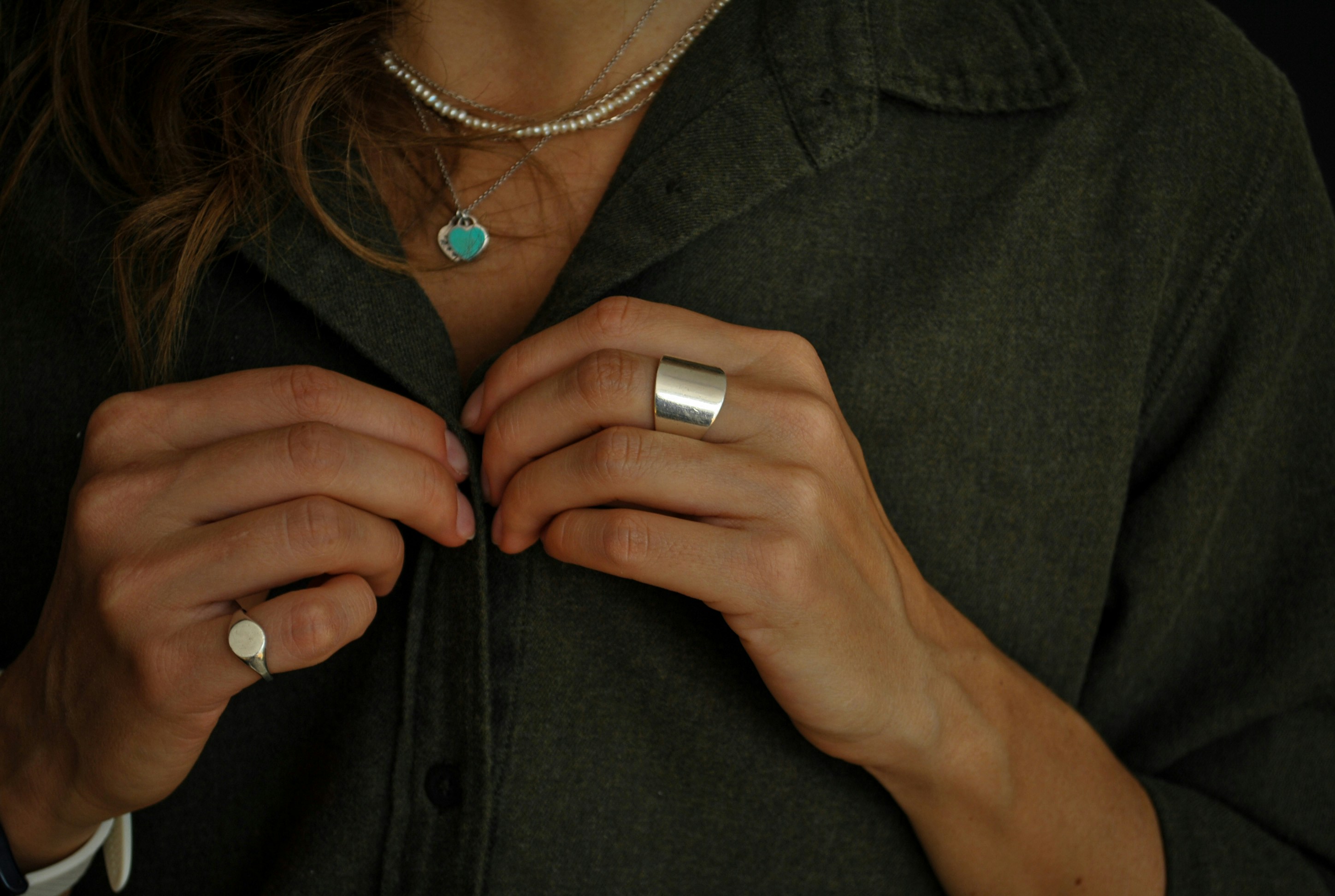 Close-up of a person's hands adjusting a shirt, showcasing delicate jewelry including rings and a necklace. The focus is on the intricate details of the accessories.