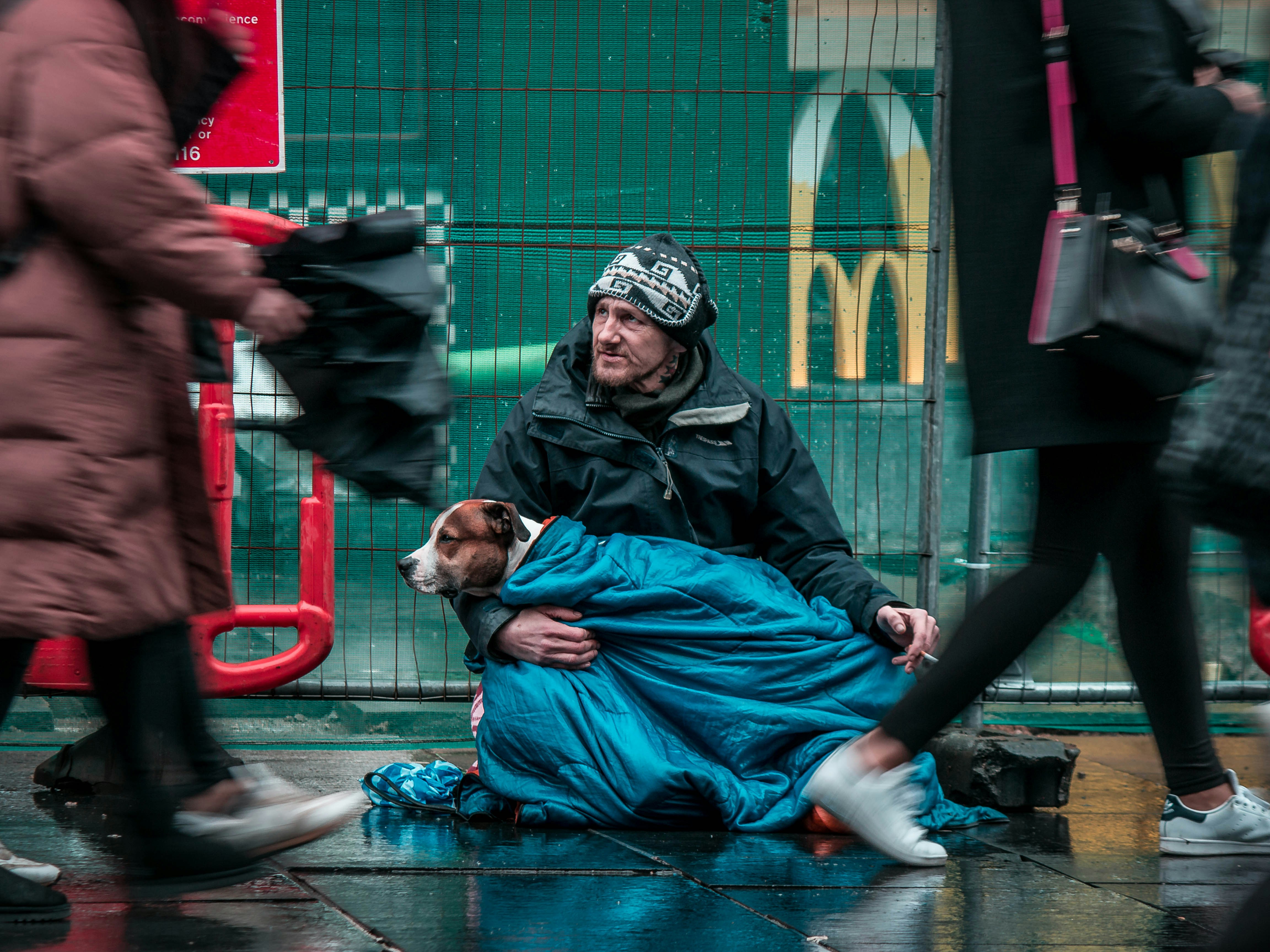A person sitting on wet pavement with a dog, wrapped in a blue blanket, surrounded by blurred passersby.