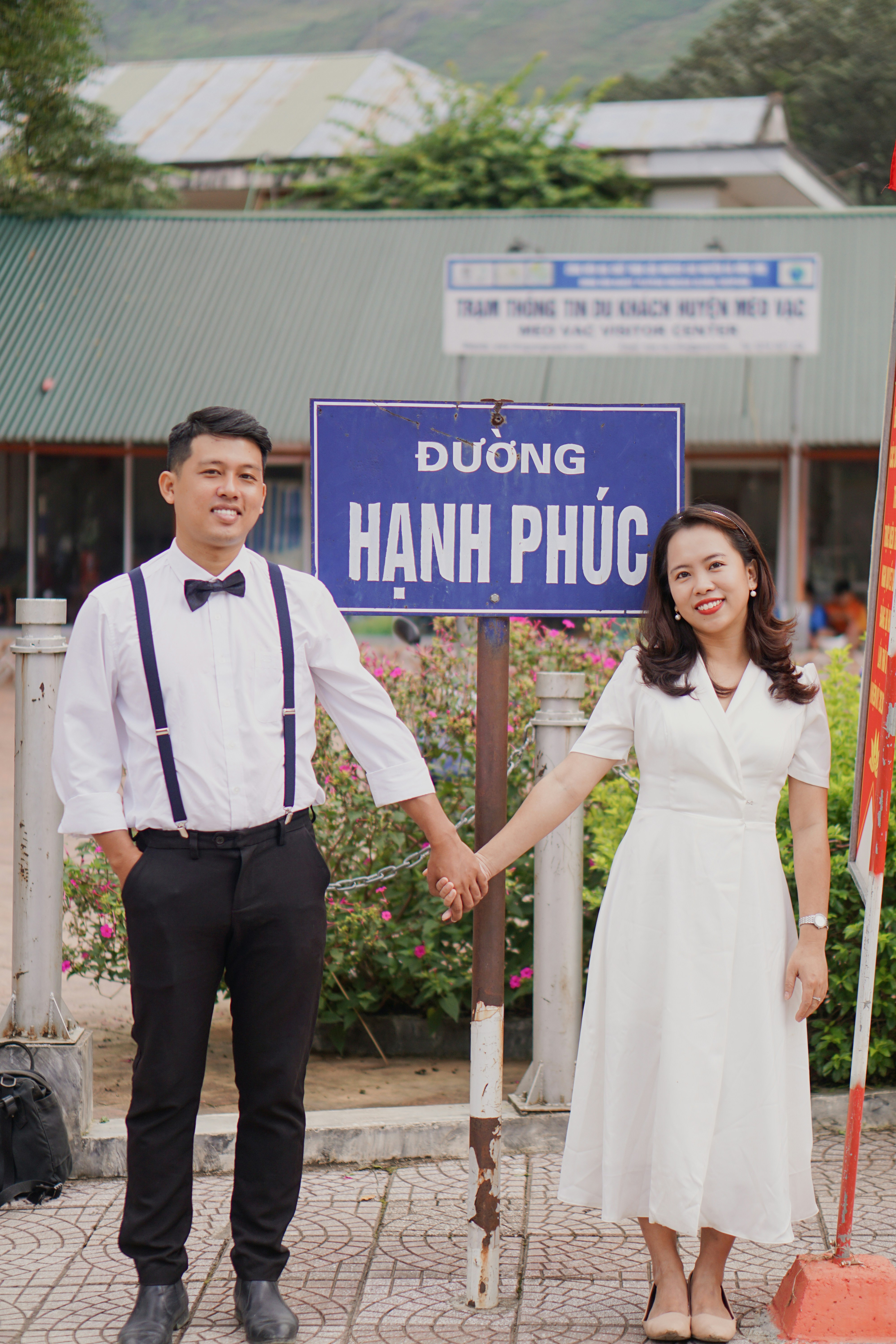Couple in formal attire holding hands beside a sign reading 'Hanh Phuc' with a visitor center in the background.