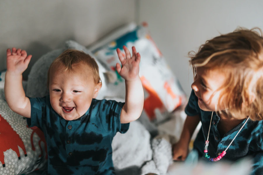 Two young children in blue outfits share a joyful moment on a cozy bed with colorful cushions.