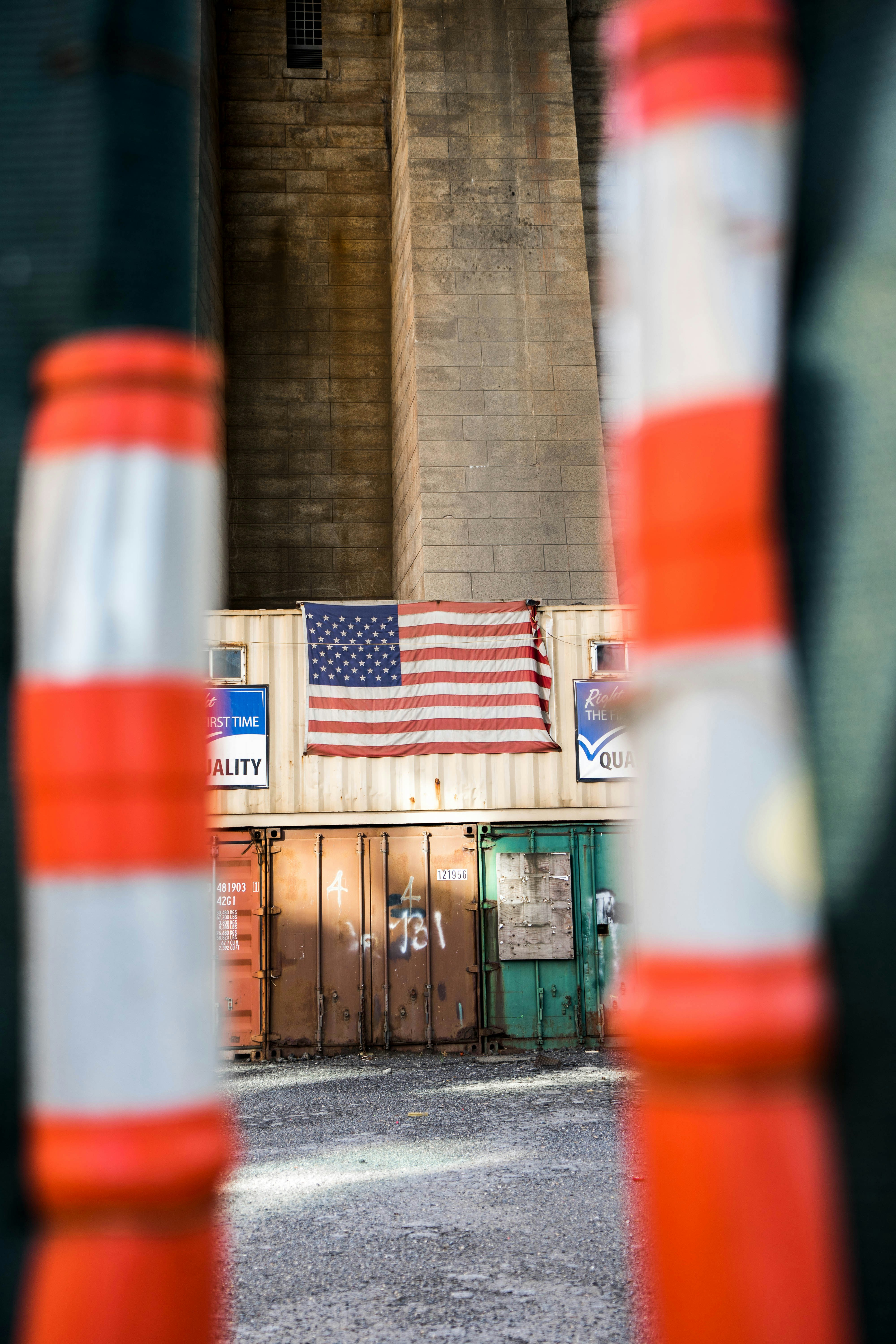 An American flag drapes over a shipping container, framed by construction cones, capturing the intersection of urban life and patriotism.