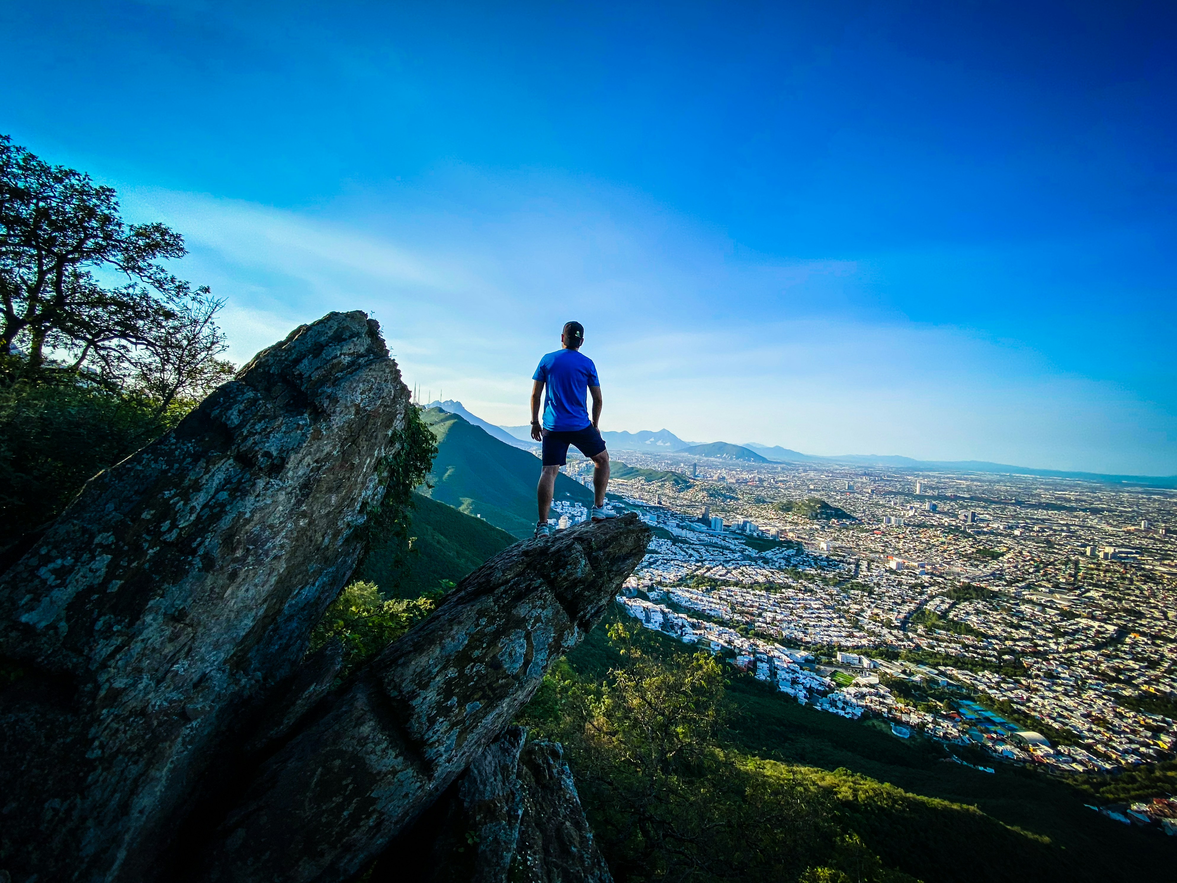 Person stands on rocky peak overlooking expansive city under a clear blue sky.
