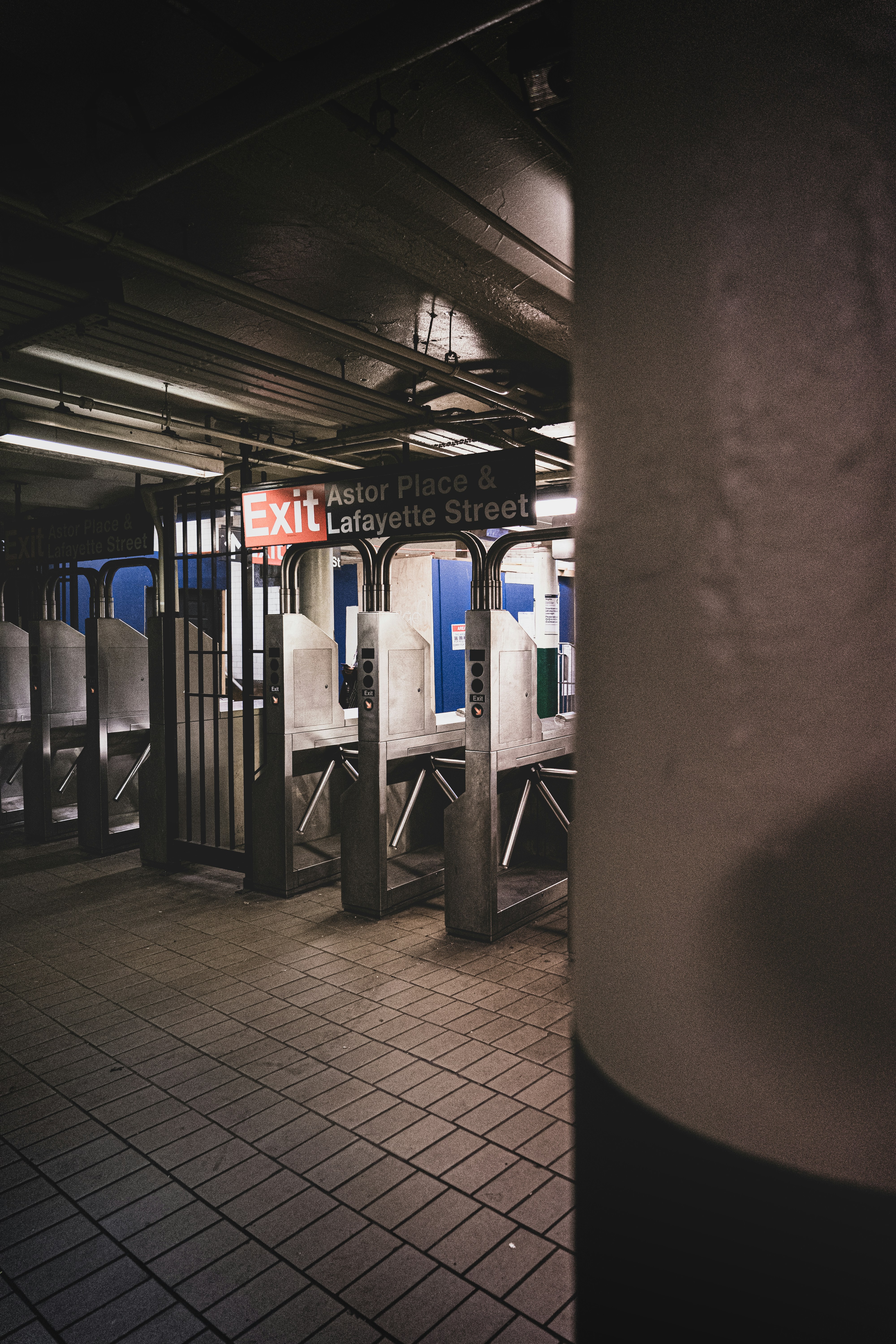 Exit sign at Astor Place & Lafayette Street in a subway station, framed by turnstiles and industrial architecture.