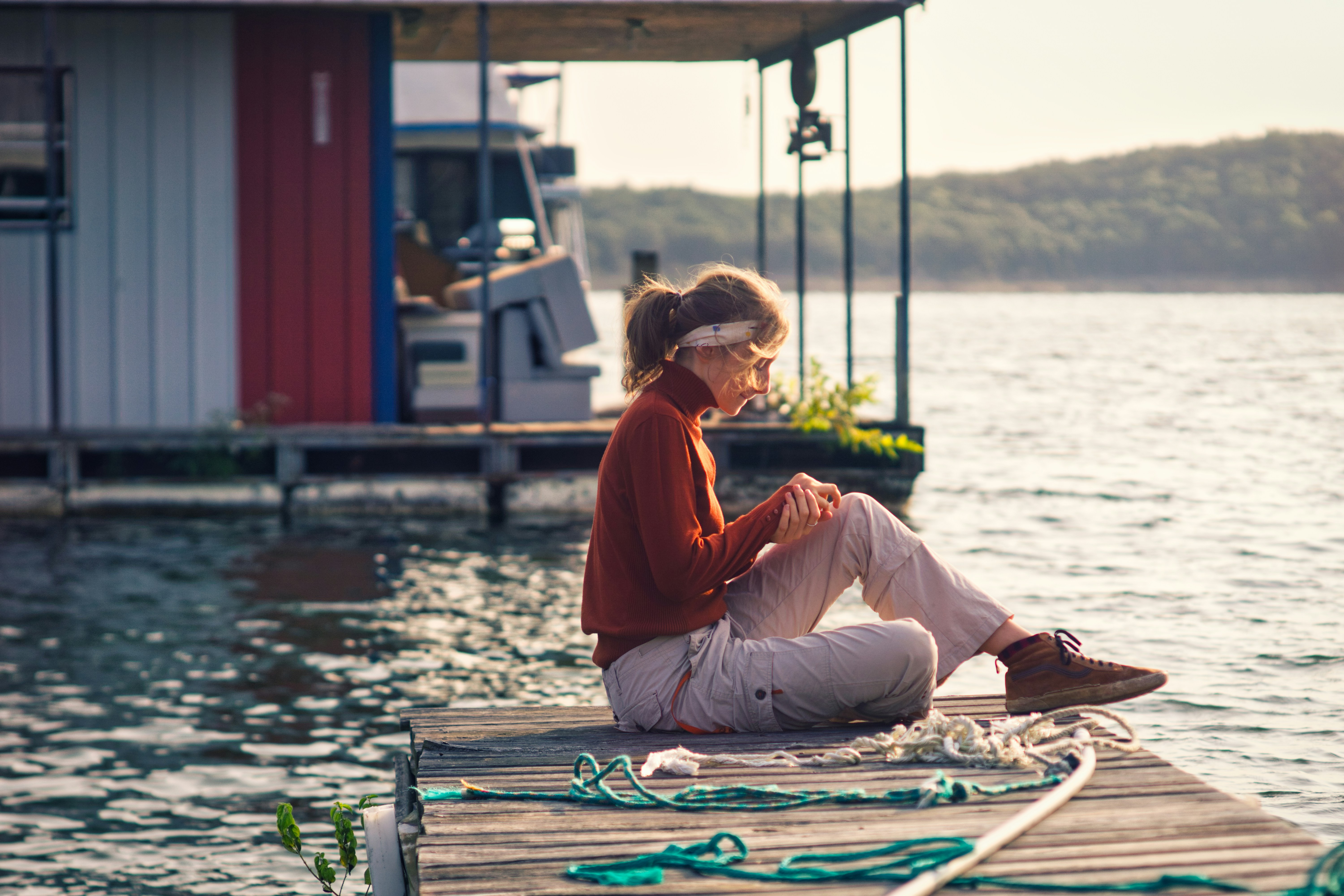 A woman sits thoughtfully on a dock, surrounded by serene water and a colorful boathouse. The gentle light enhances the tranquil atmosphere.