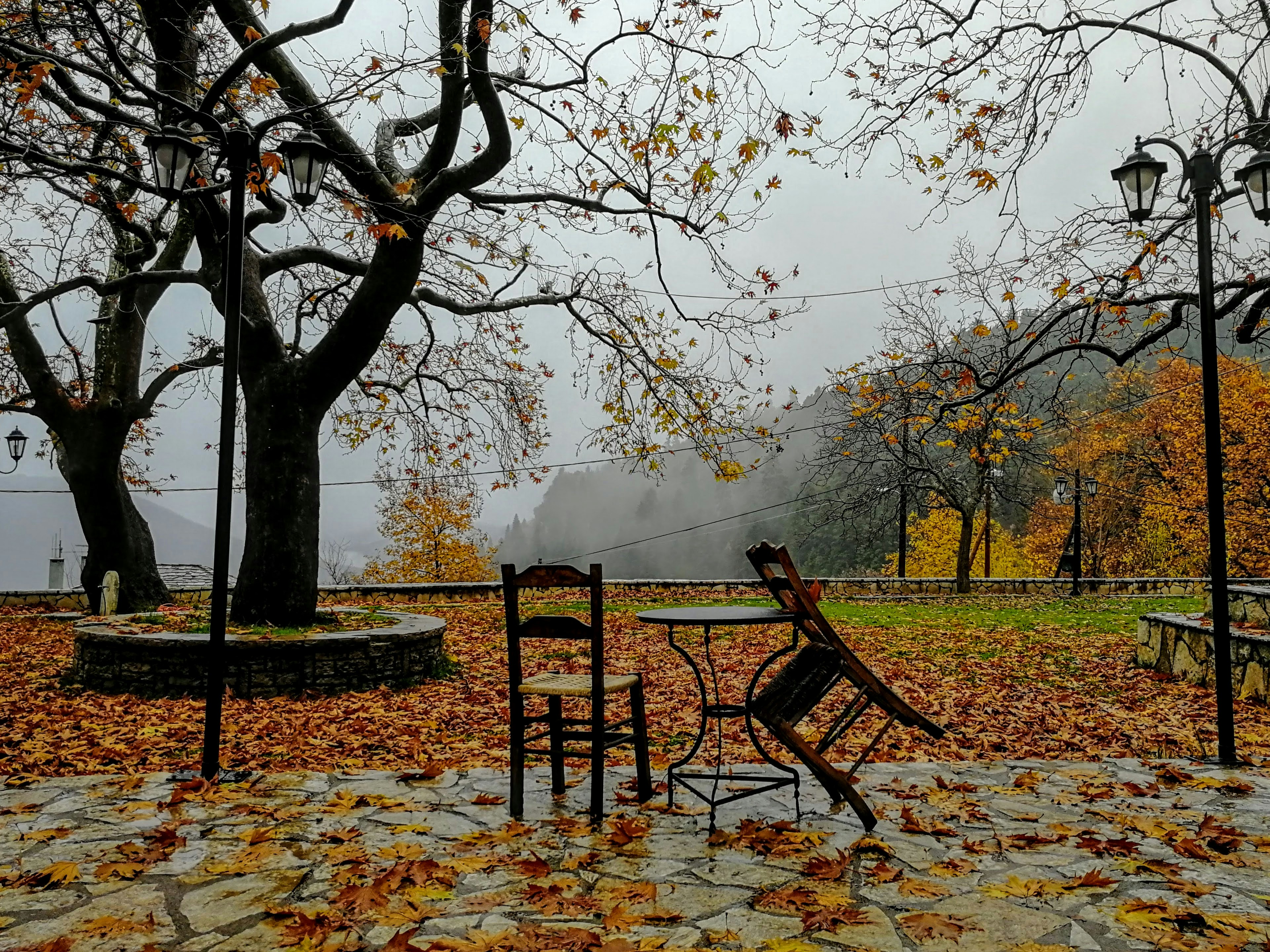 Autumn park scene with leaf-covered ground, empty chairs around a small table on a stone-paved area, and misty hills in the background.