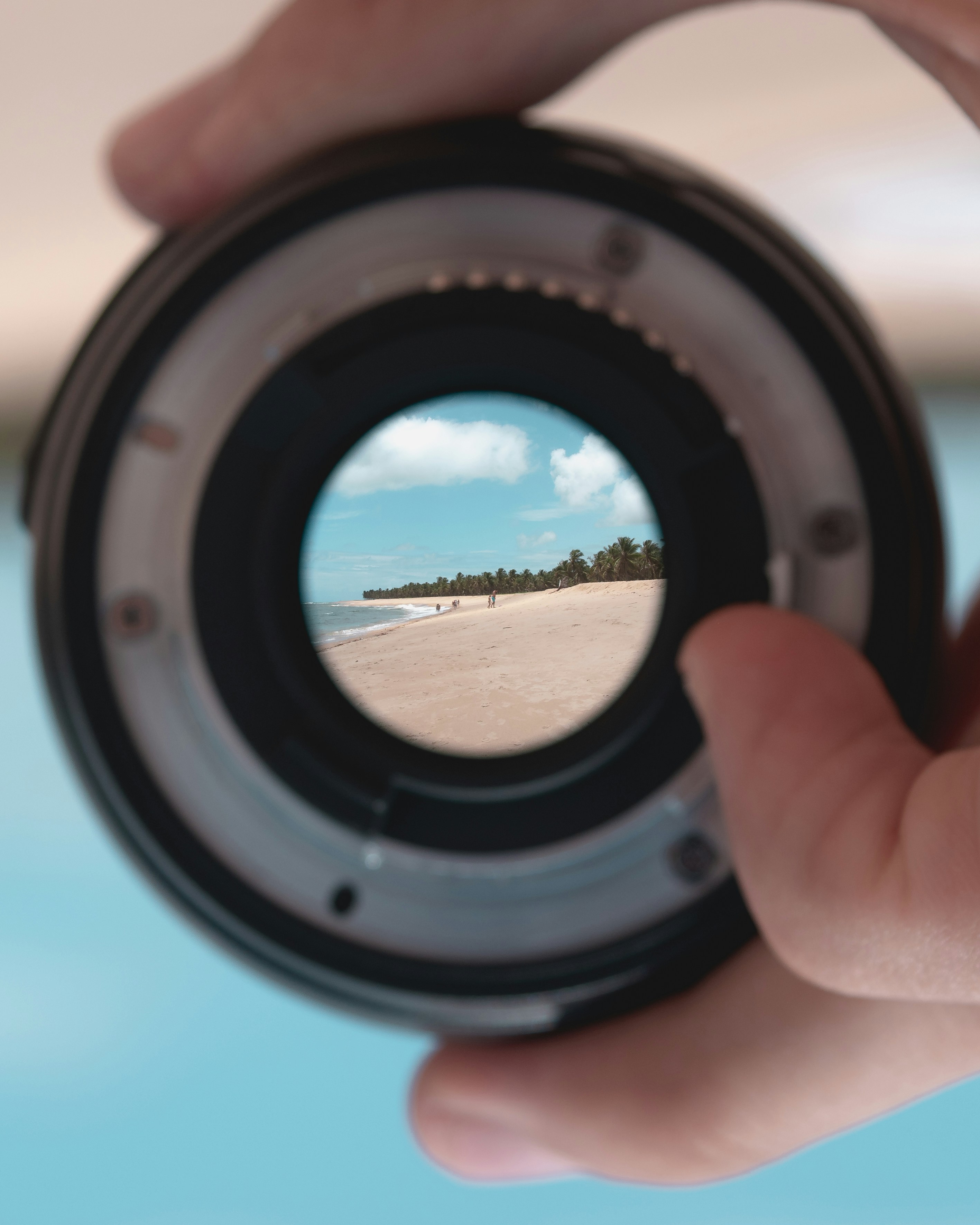 A hand holds a camera lens, framing a serene beach scene with gentle waves and palm trees under a partly cloudy sky.