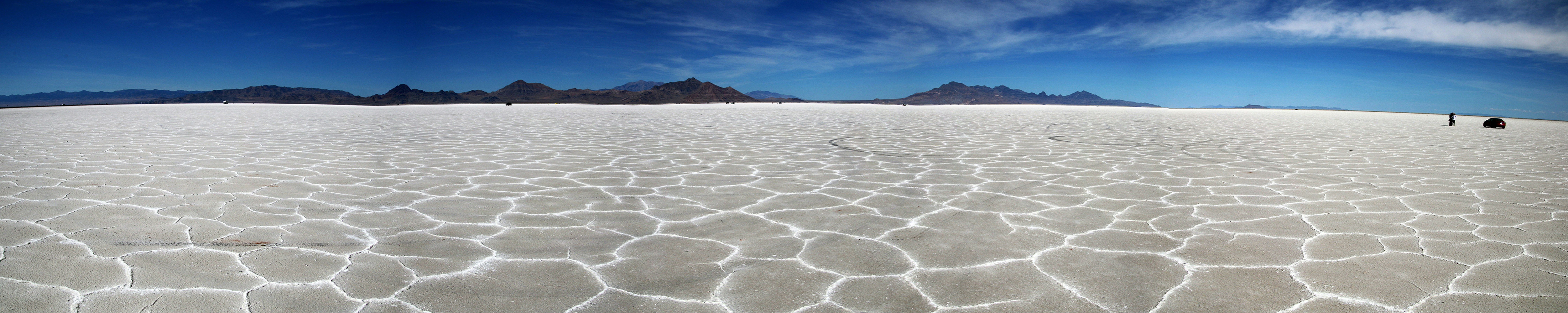 Hexagonal salt formations stretch towards distant mountain ranges under a bright blue sky.