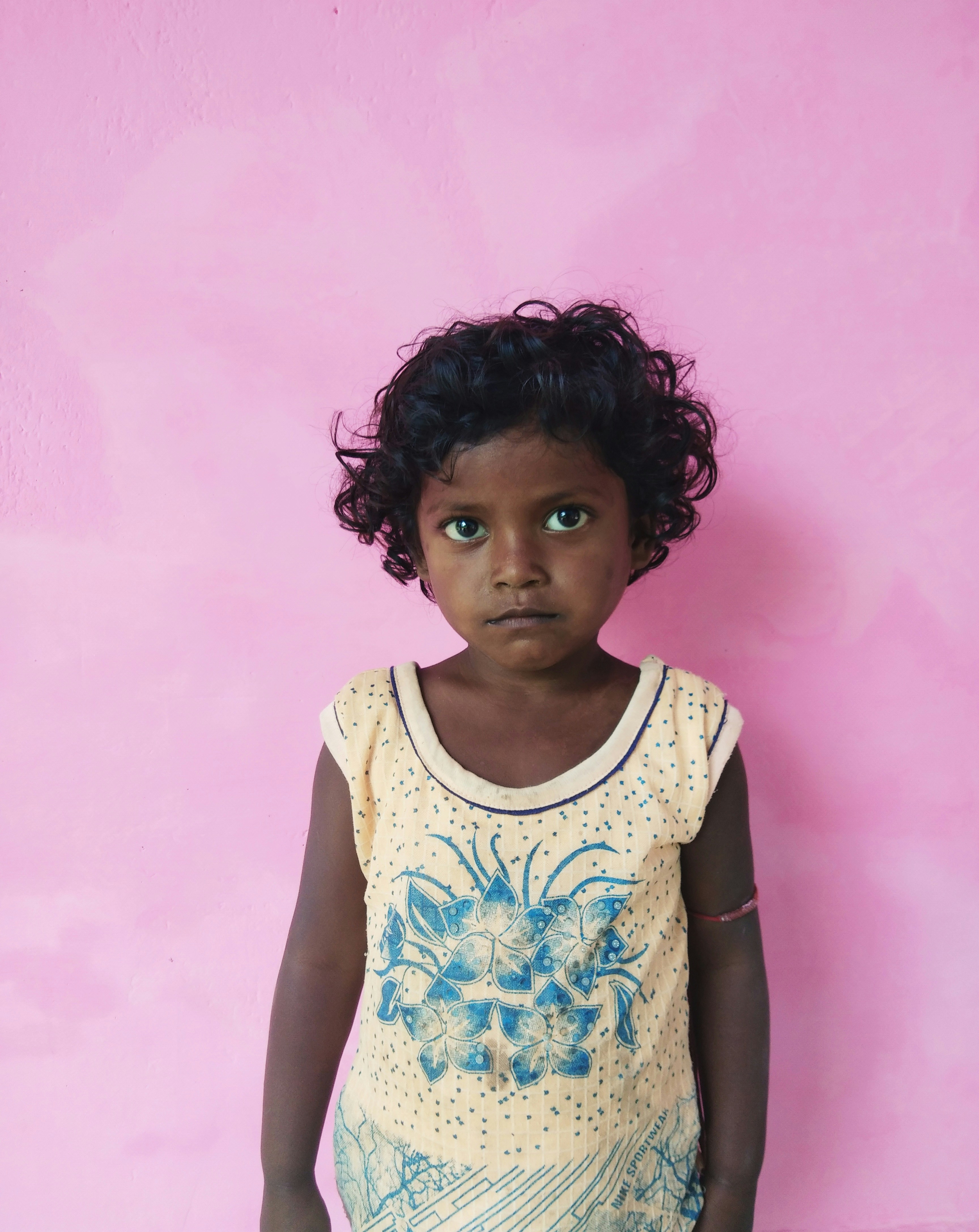 Portrait of a young girl with curly hair standing against a pink wall, wearing a pale yellow sleeveless top with a blue graphic.