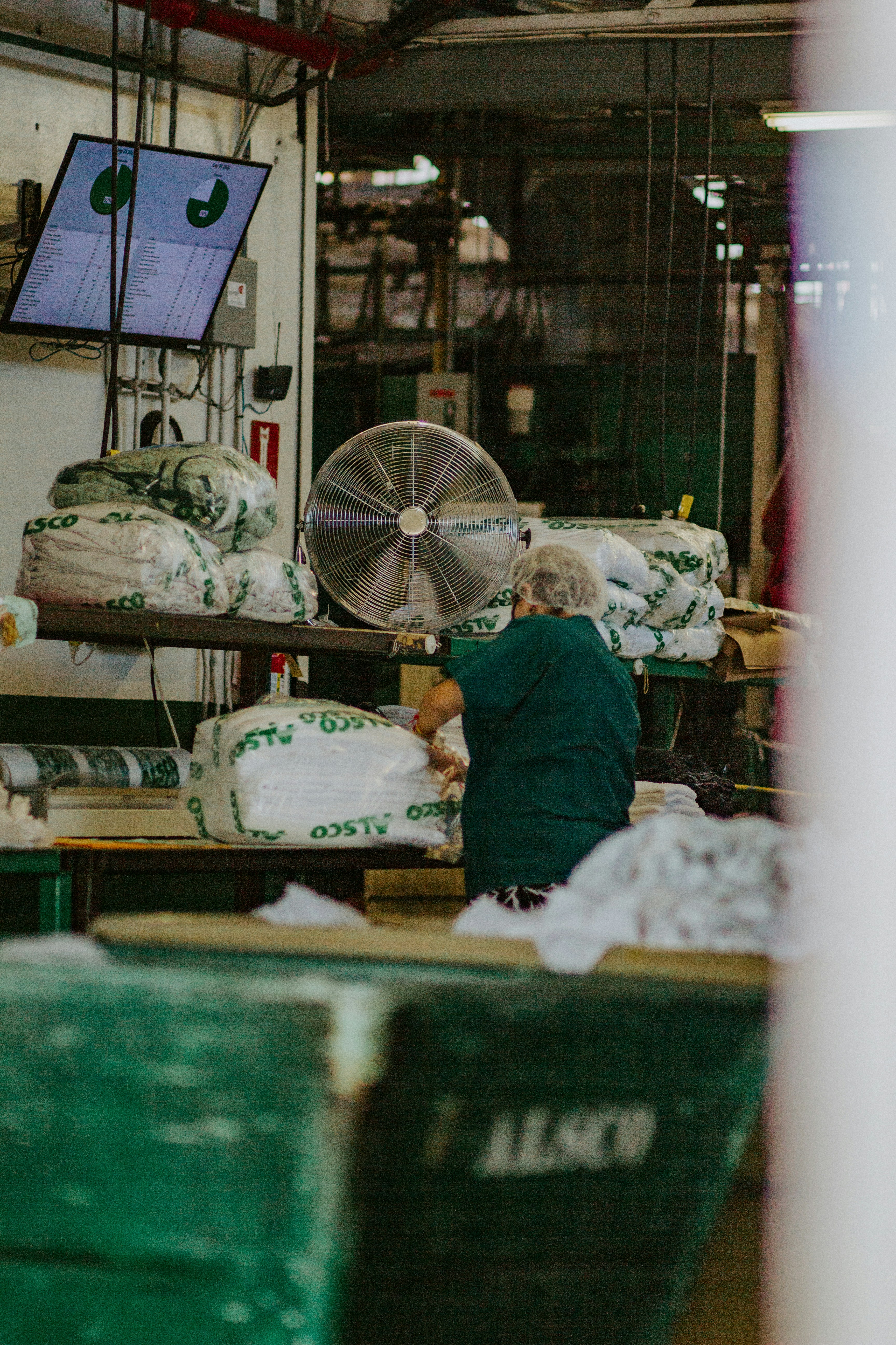 A warehouse worker in a green uniform and hairnet sorts large bags at a cluttered table, with a large oscillating fan behind. This candid photograph captures routine industrial labor amid stacked sacks and machinery.