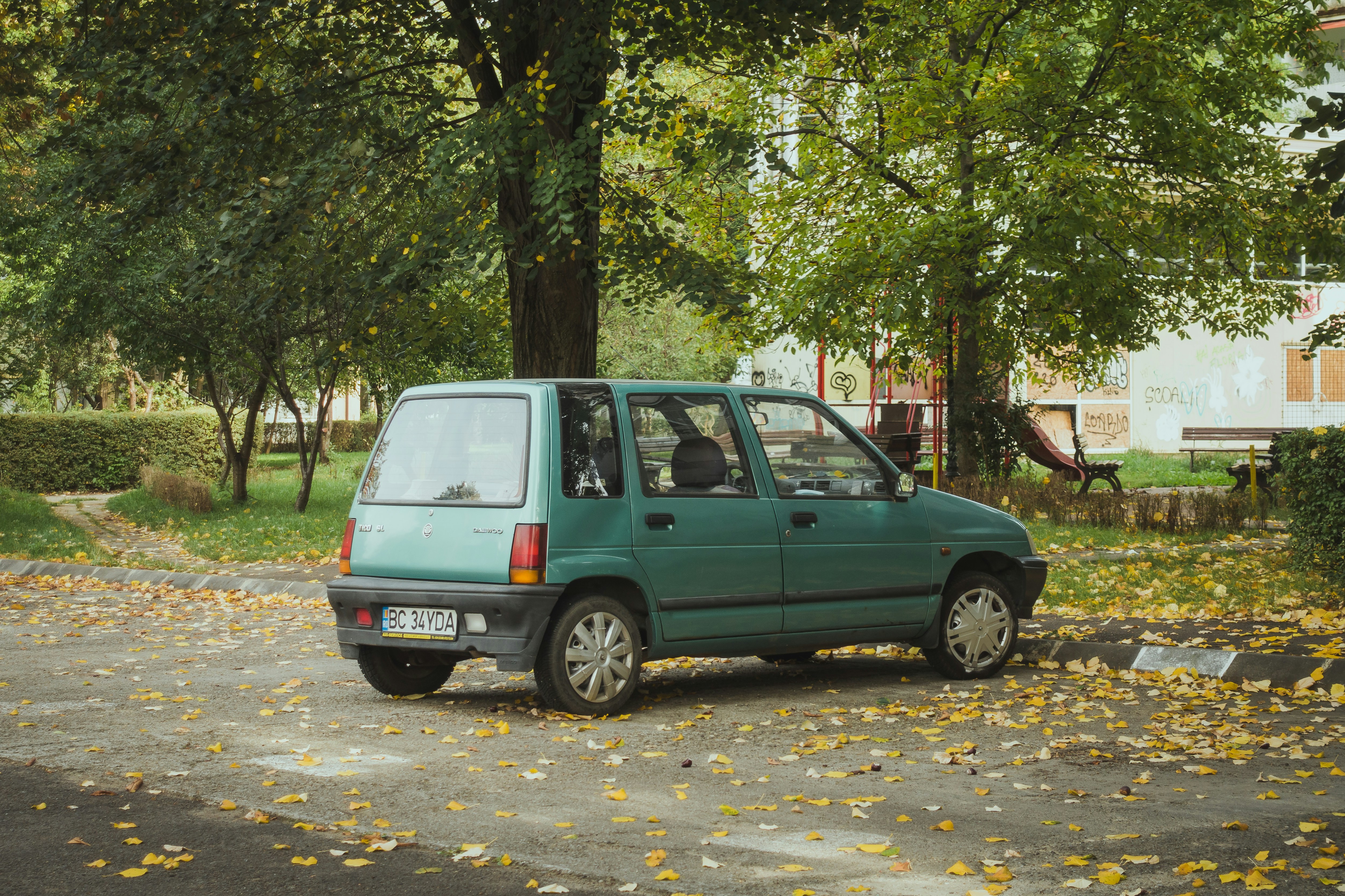Teal hatchback parked under lush green trees on a leaf-strewn street in autumn.