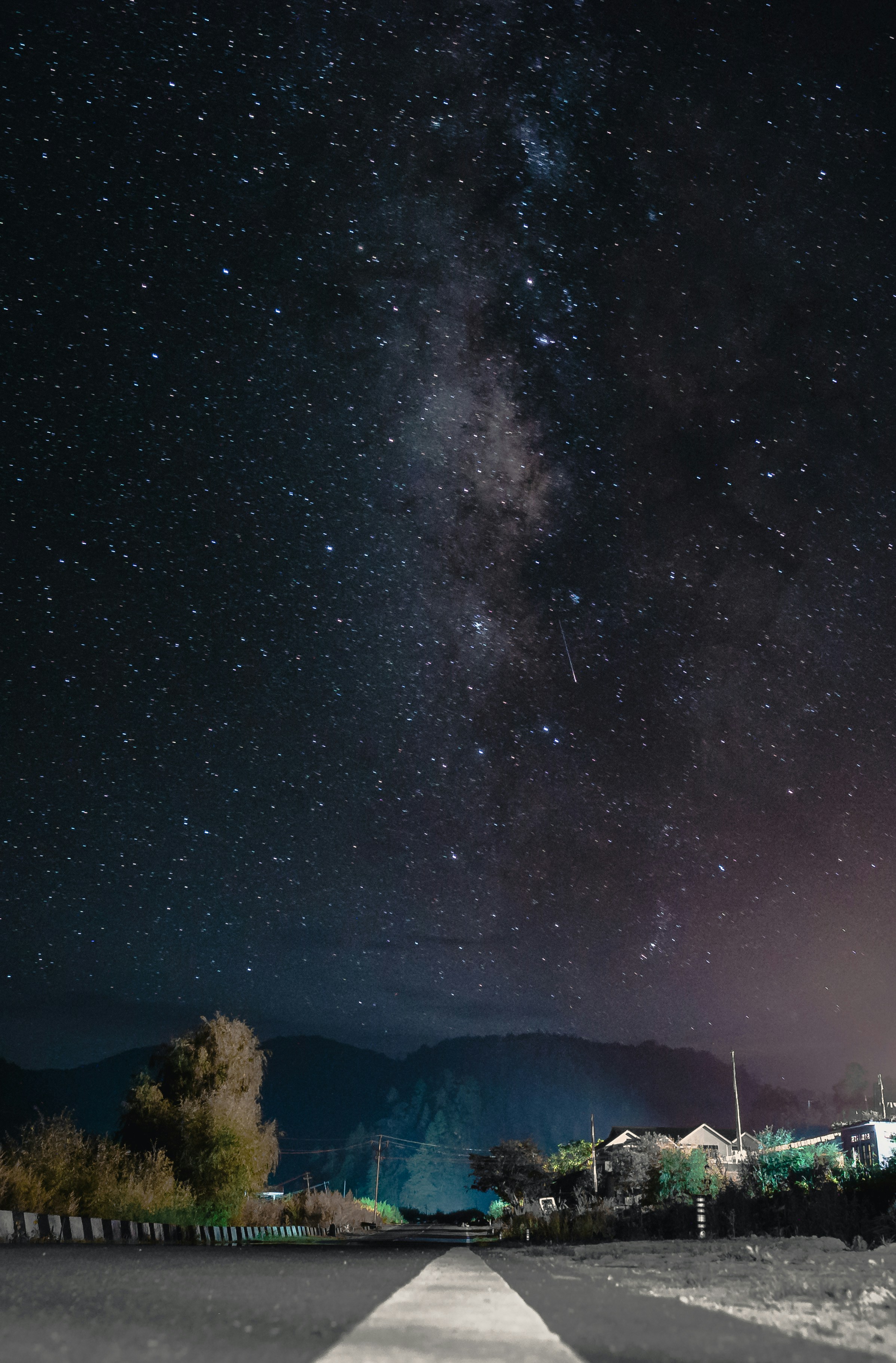 Milky Way stretching across the night sky above a quiet road, framed by distant mountains and subtle vegetation. A faint shooting star adds to the cosmic scene.