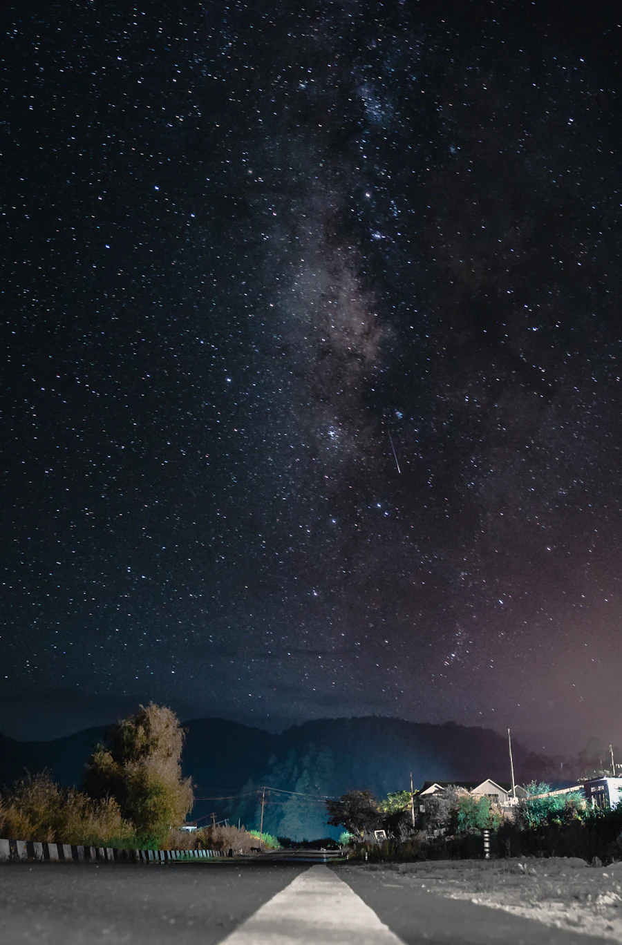 A star-filled night sky with the Milky Way arcing above a dark mountain road and small houses at the horizon. A faint streak of light cuts through the sky near the center.