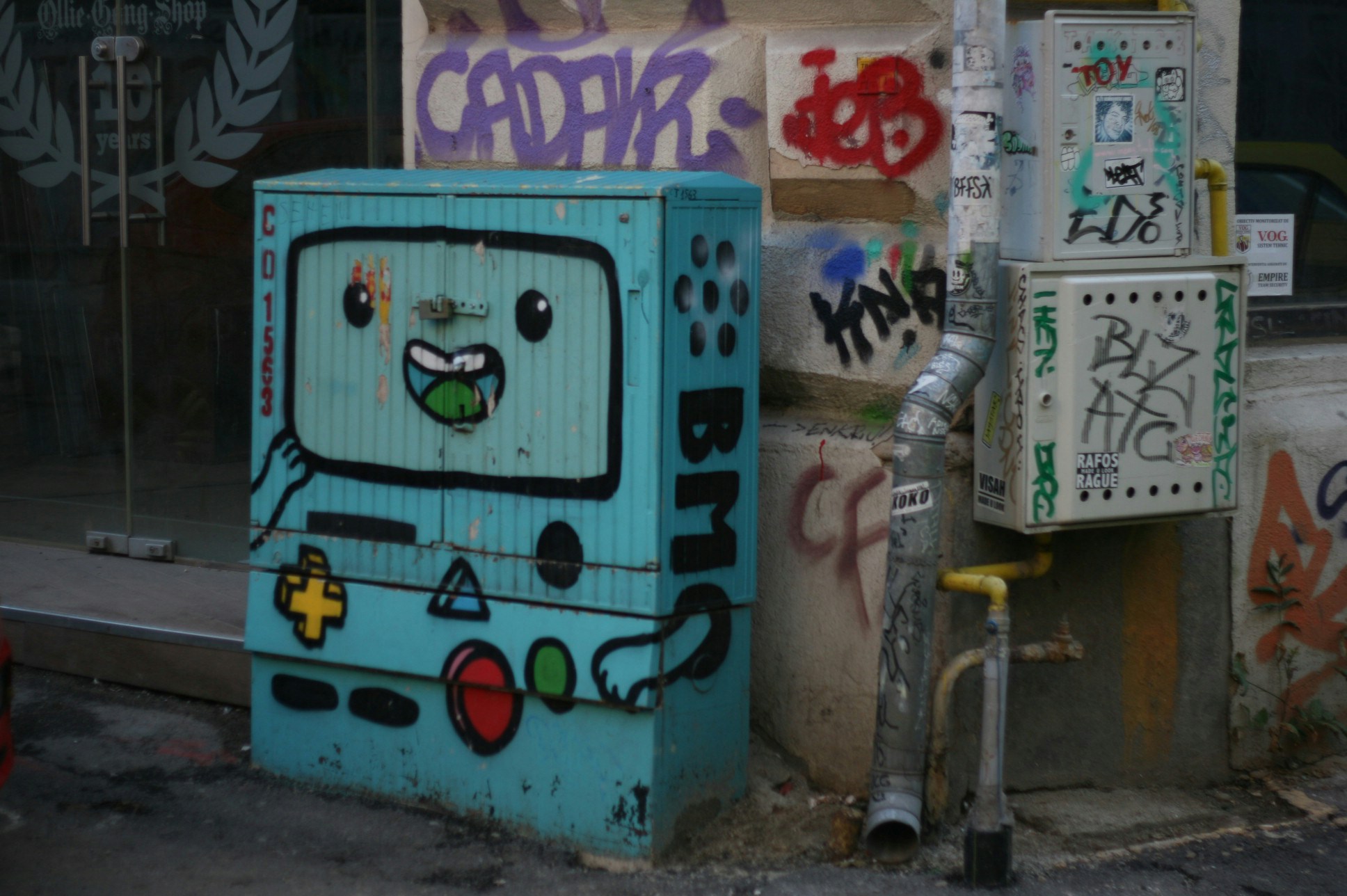 Cartoon-style graffiti of a game console character on an electrical box surrounded by urban tags.