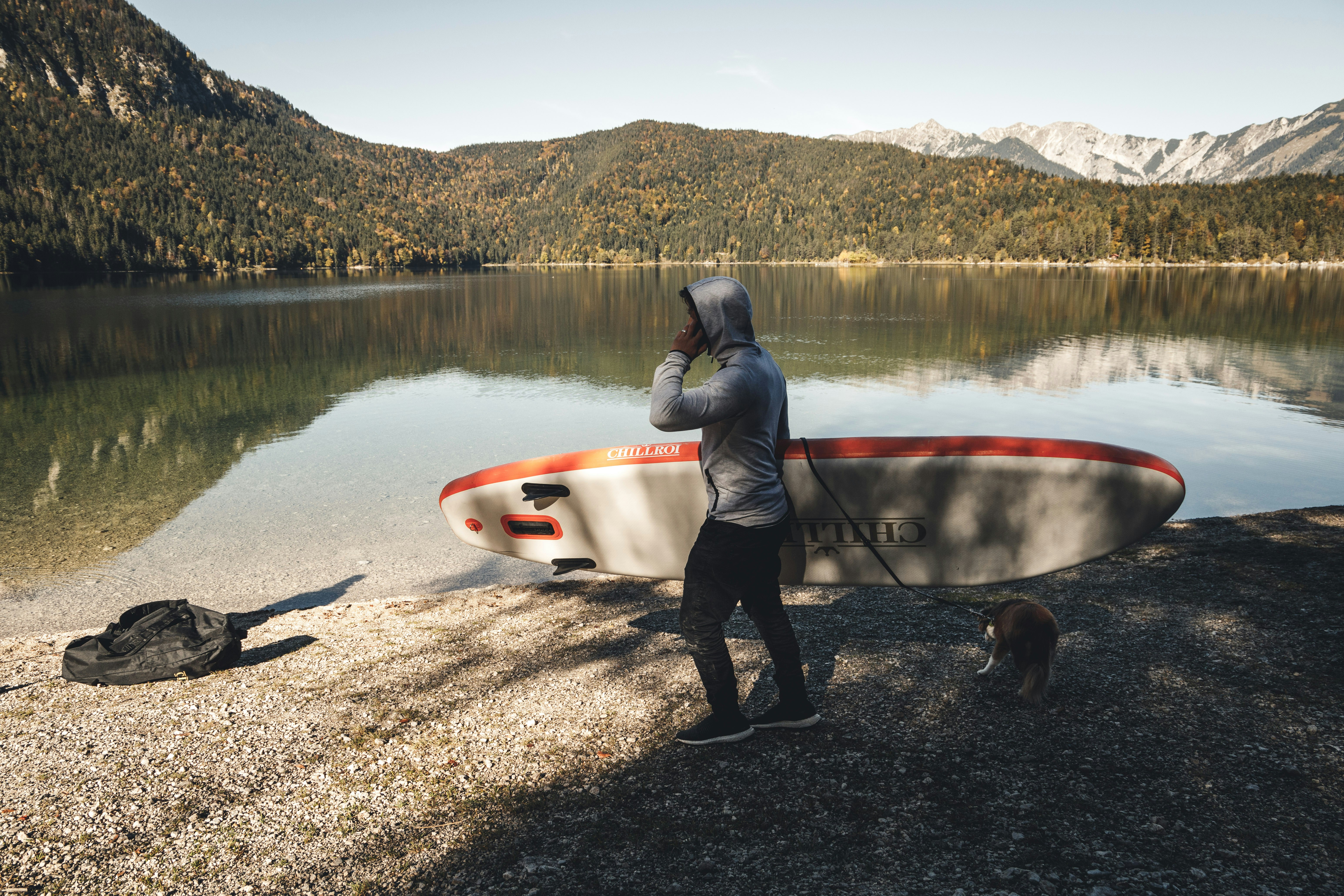 Person walking along the shore with a paddleboard and a dog, surrounded by a tranquil lake and mountains in the background.