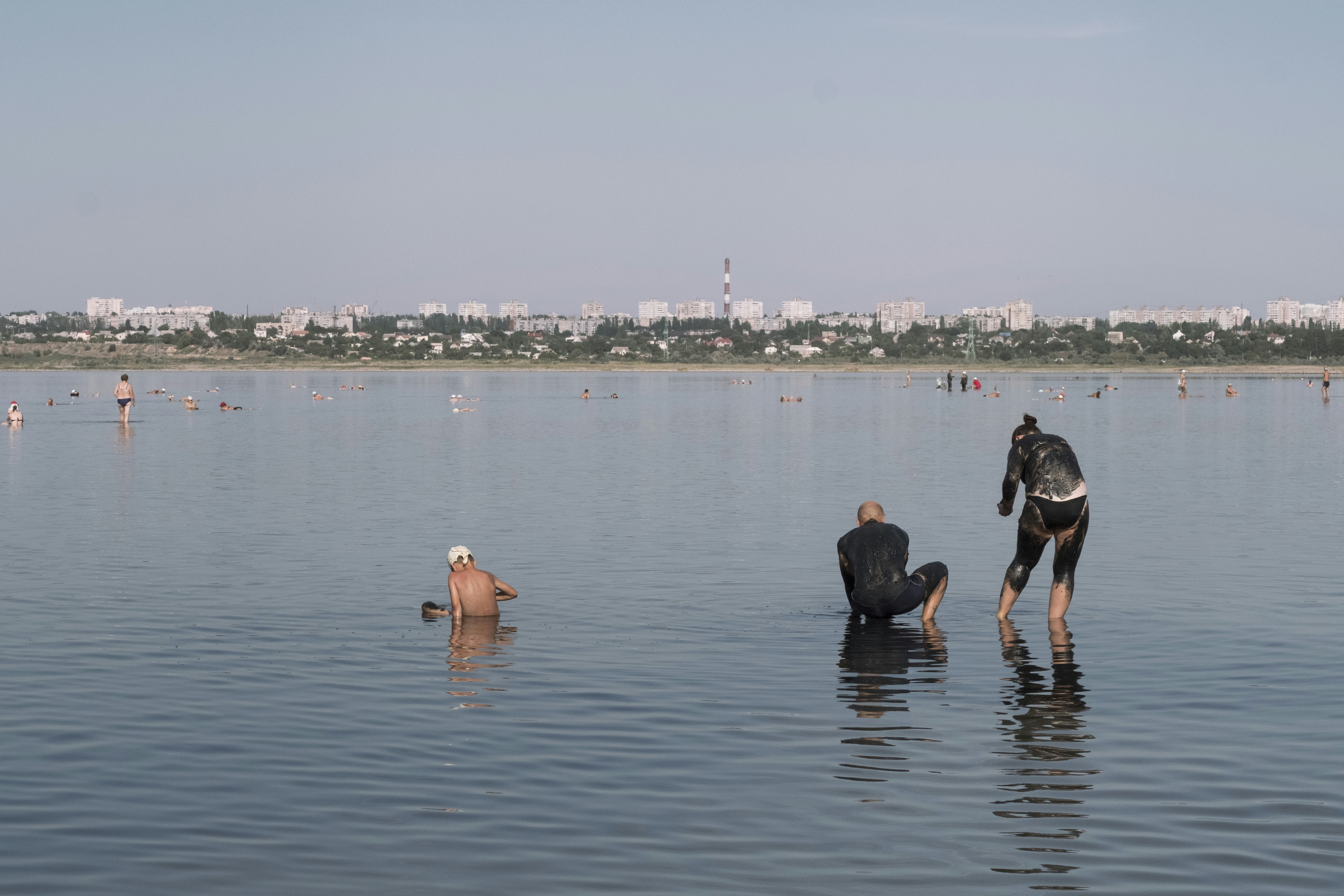 Two men wade in a tranquil body of water, while others enjoy the coolness in the background. The city skyline looms softly in the distance.