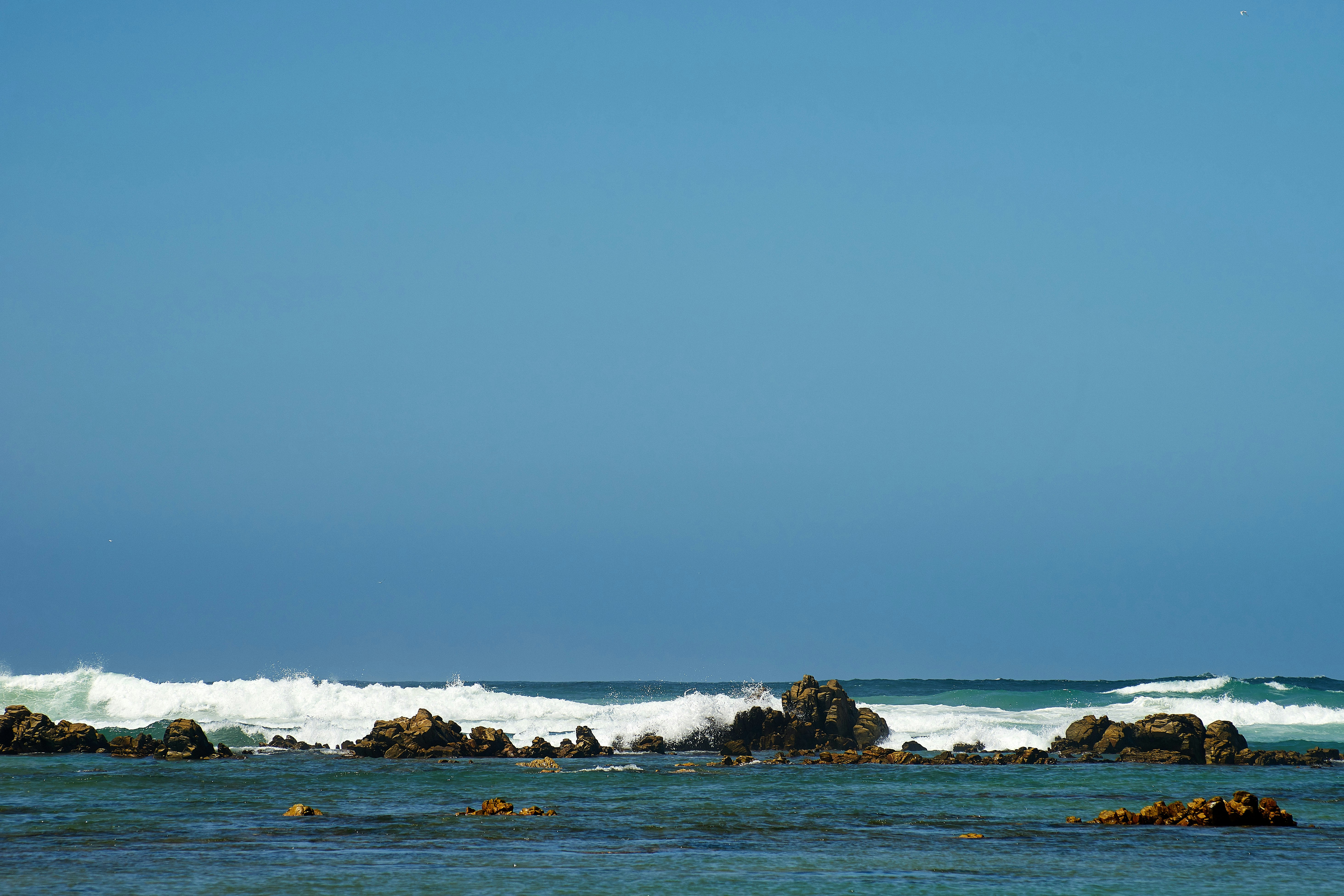 Waves crashing against rocky outcrops under a clear blue sky, capturing the serene yet powerful essence of coastal waters.