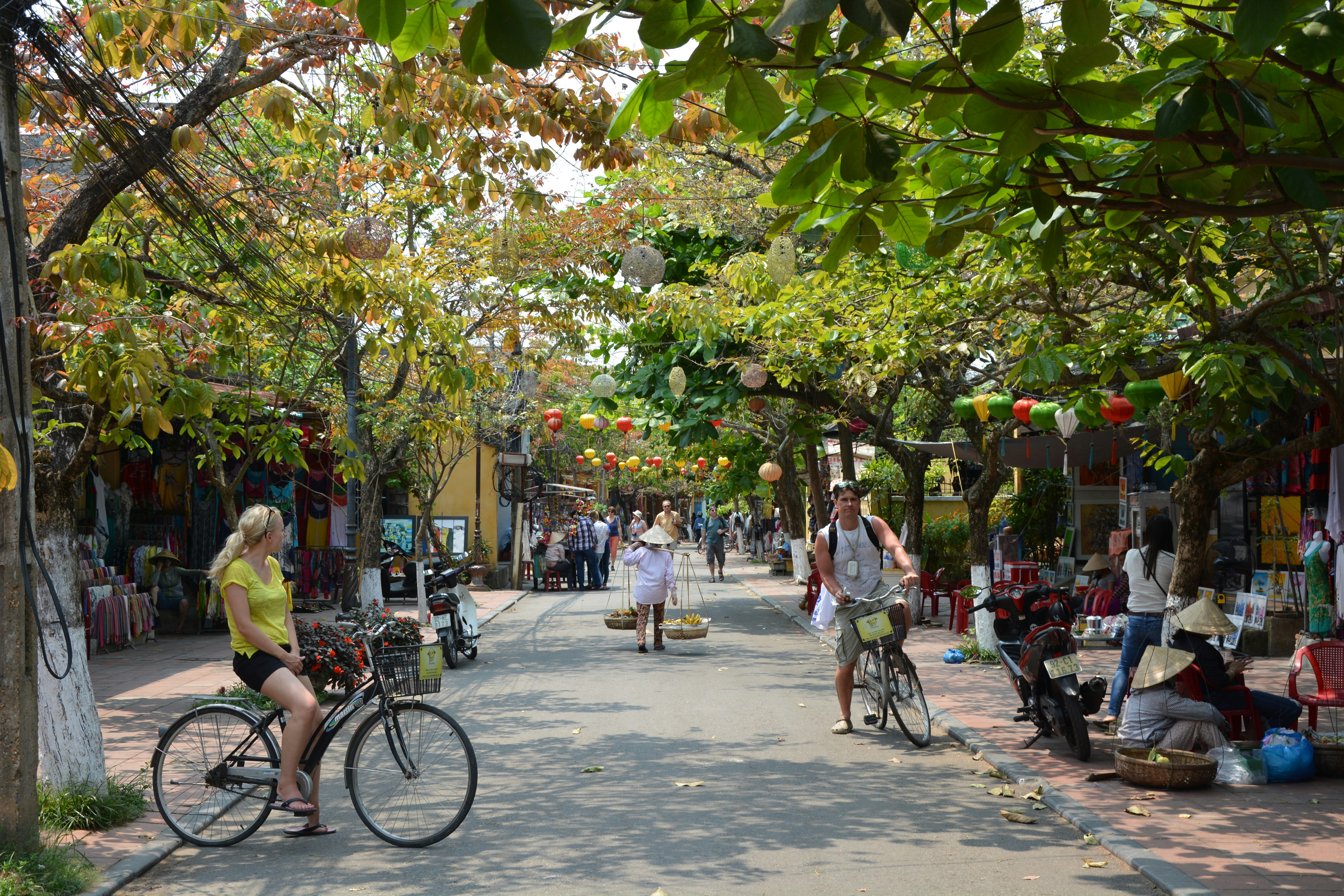 Tree-lined street with colorful lanterns and people on bicycles in Hoi An, Vietnam.