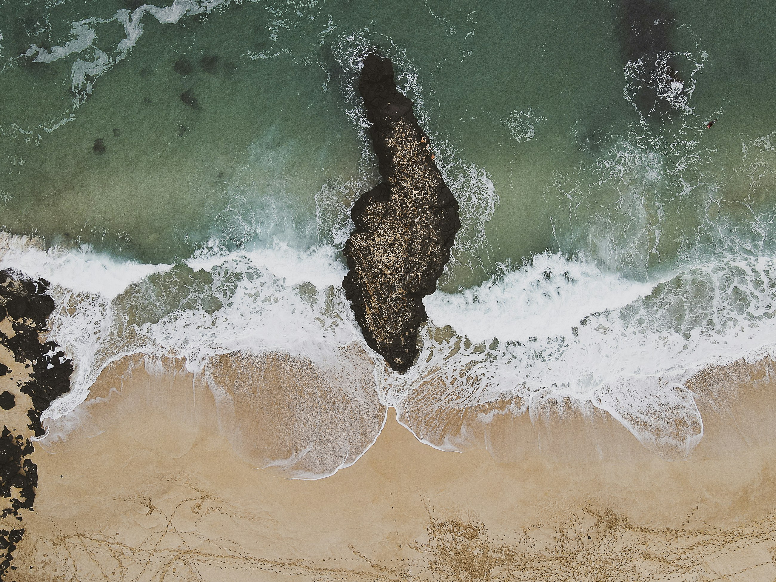Aerial view of a rocky outcrop surrounded by crashing waves on a sandy beach, highlighting the contrast between land and sea.