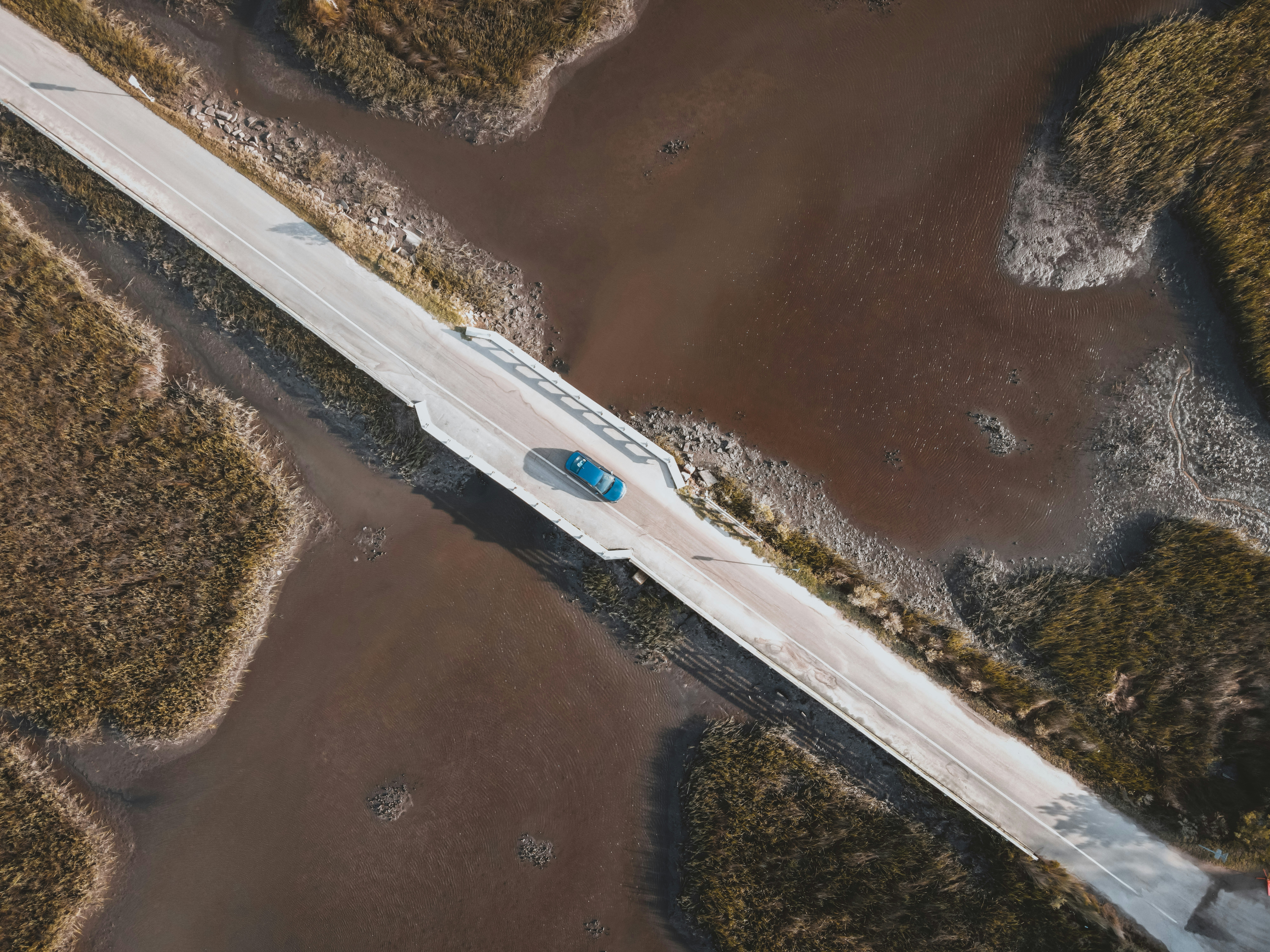 Car crossing a narrow bridge over marshy waters with surrounding grassy patches.