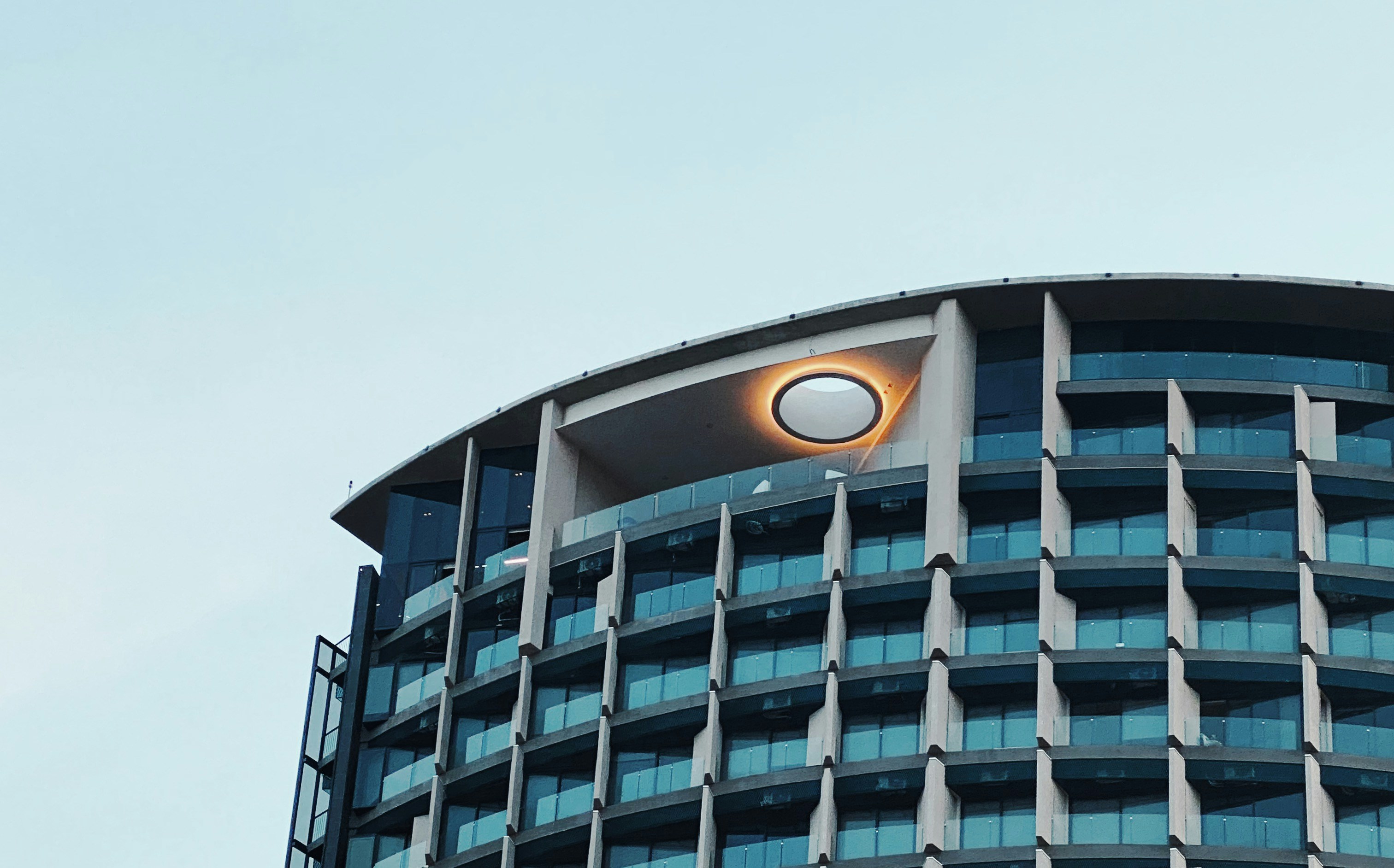 Curved building facade with symmetrical windows and a circular design element glowing against a pale blue sky.