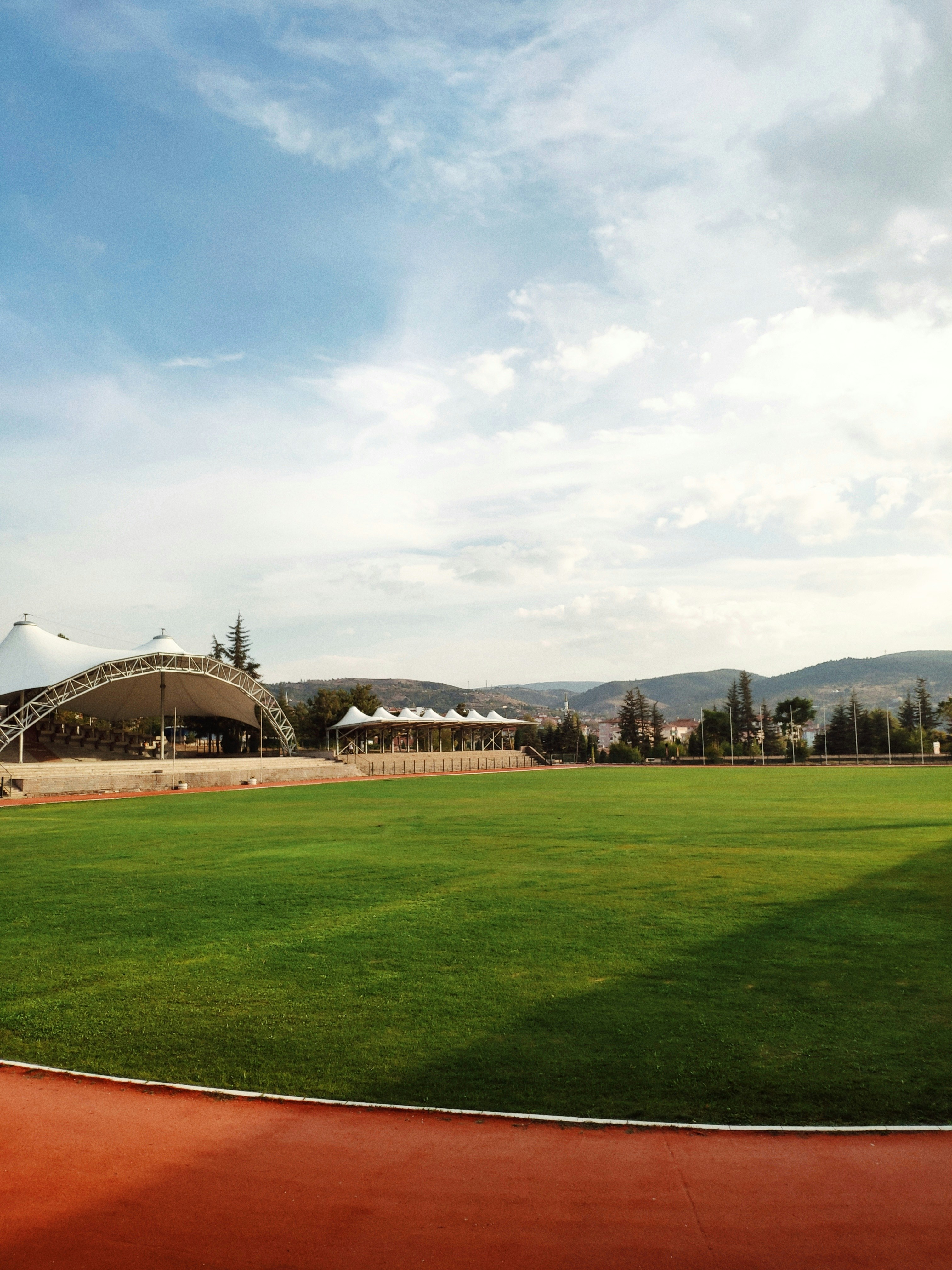 Vibrant green athletic field framed by modern structures and distant hills under a partly cloudy sky.
