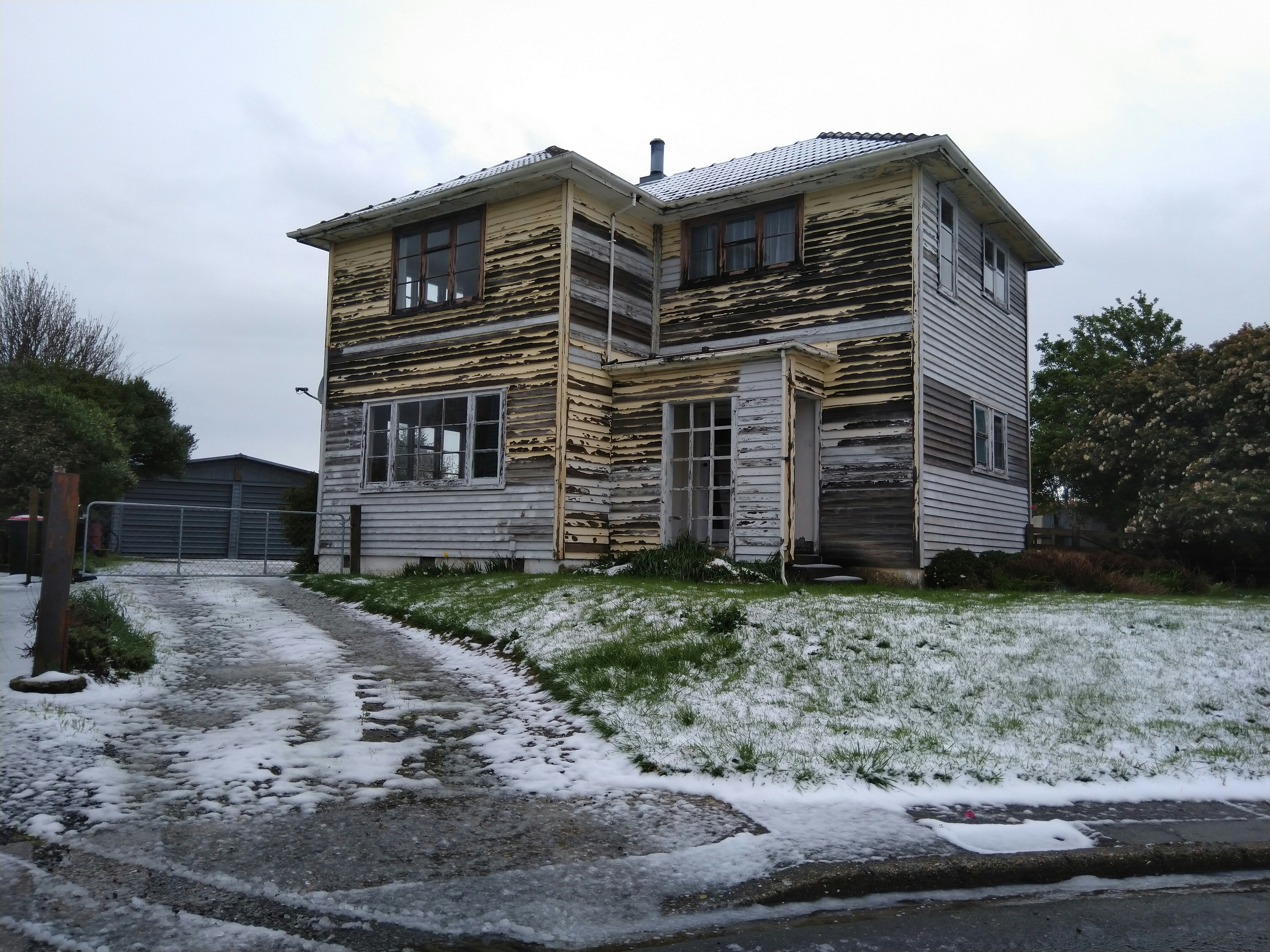 Two-story house with extensive burn damage to the upper level, exposed charred wood, and a snow-dusted front yard. Overcast daylight emphasizes a weathered residential facade.