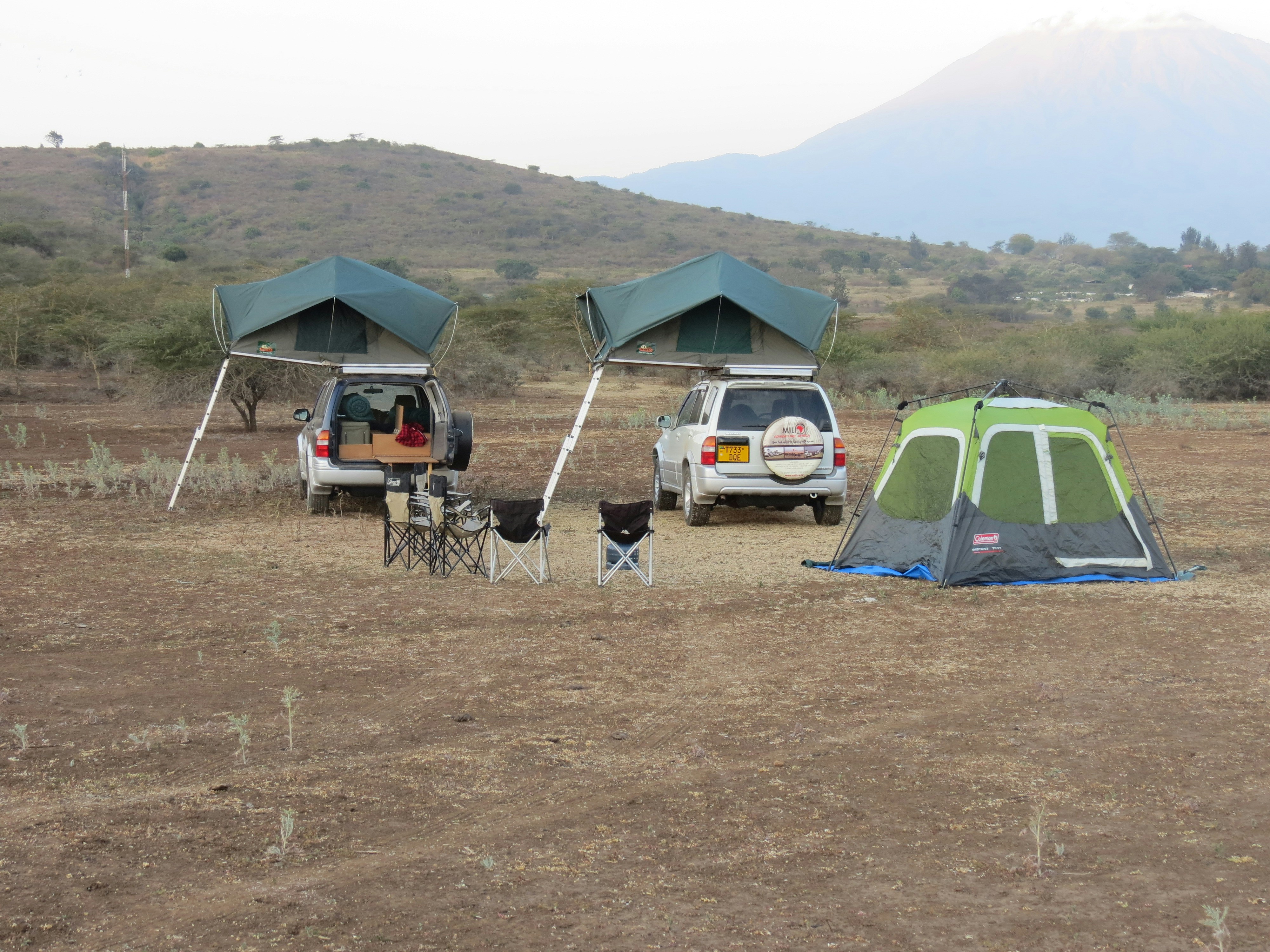 Two SUVs with rooftop tents flank a small campsite on a dry plain, with folding chairs and a green tent nearby. A pair of dogs sits between the chairs, while a distant conical mountain anchors the horizon.