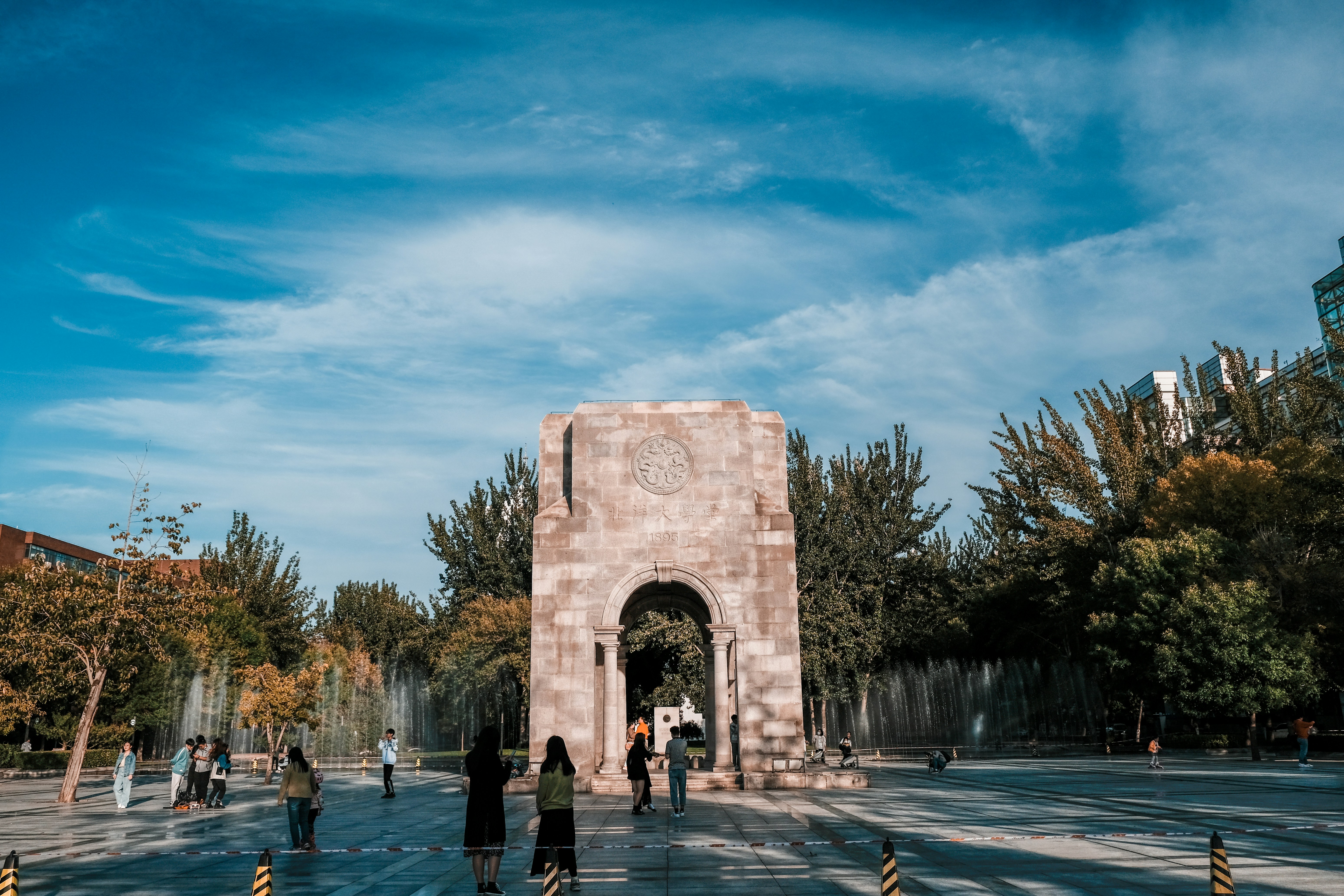 Stone archway stands amidst a tree-lined plaza under a vibrant blue sky.