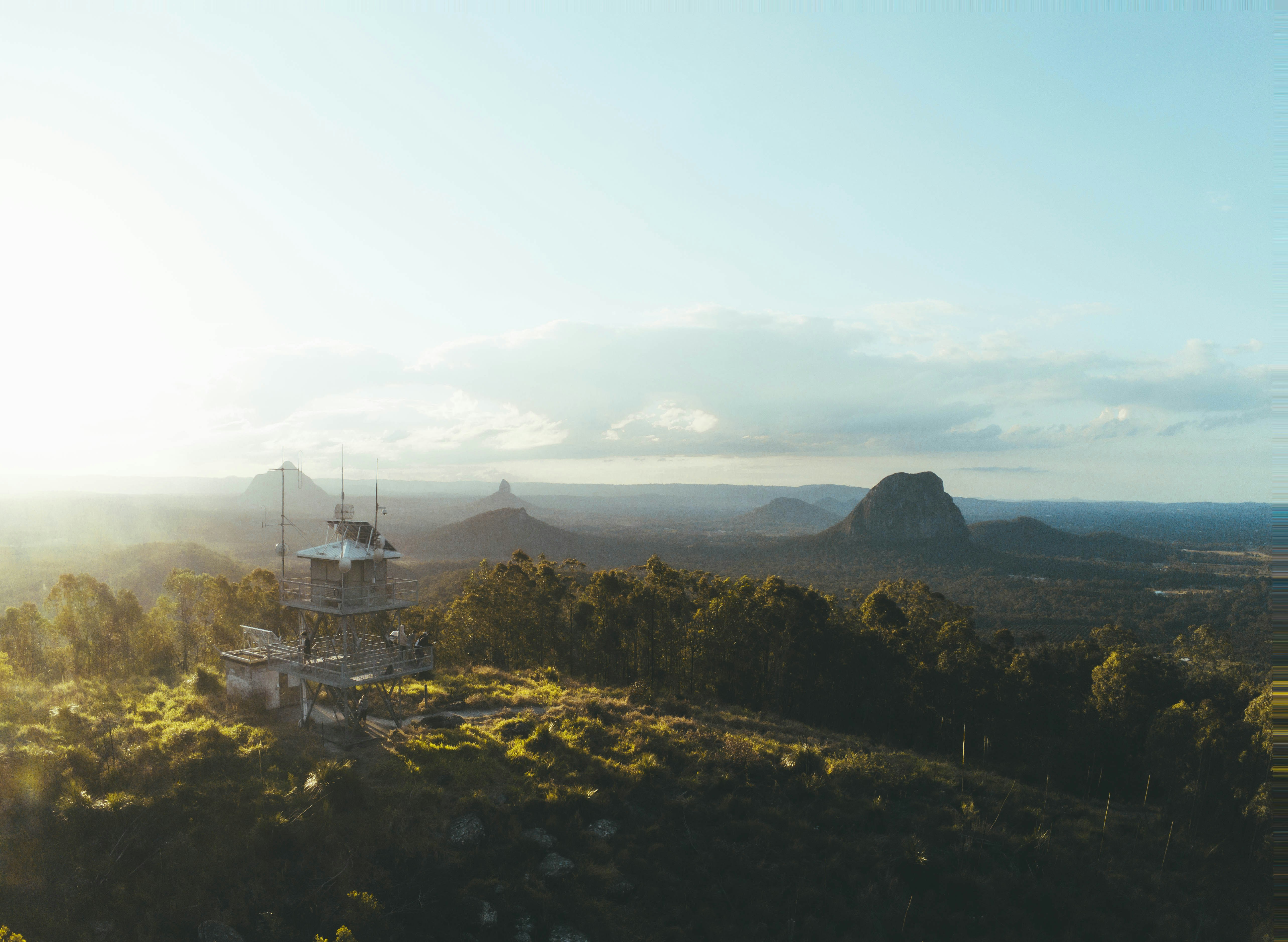 Drone-shot landscape at sunrise. A weathered radio tower sits on a grassy outcrop, with distant mesas and a softly lit horizon.