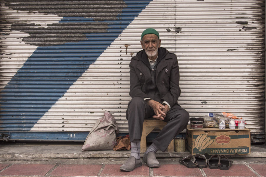 An elderly man in a green cap sits on a small stool in front of a painted corrugated metal shutter, with bags and shoes on the sidewalk beside him. The weathered wall and simple setup give the scene a quiet, urban feel.