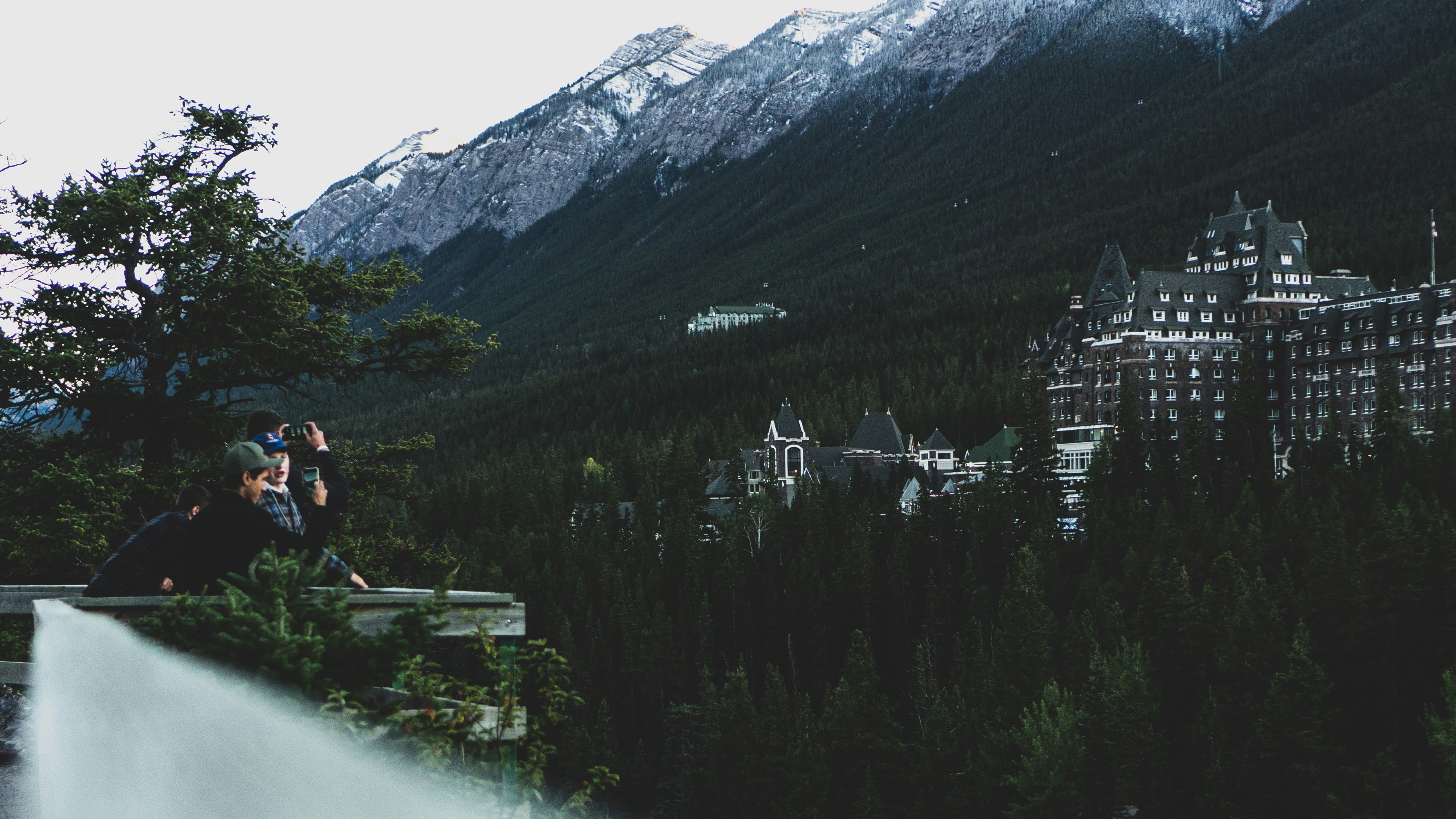 Tourists capturing the scenic view of a historic hotel amidst towering mountains and dense forests.