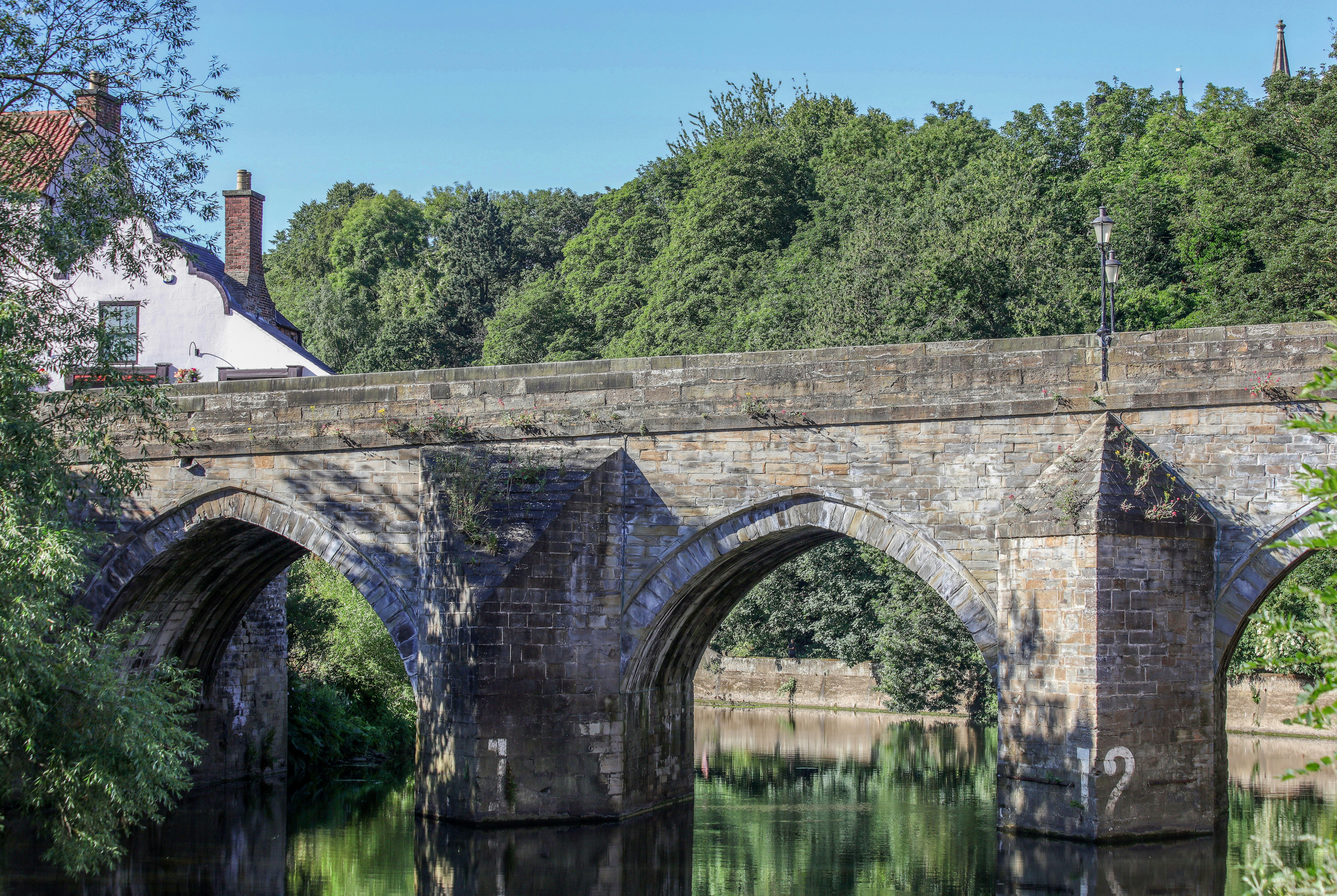 Historic stone bridge with three arches spanning a calm river, surrounded by lush greenery and a nearby house.