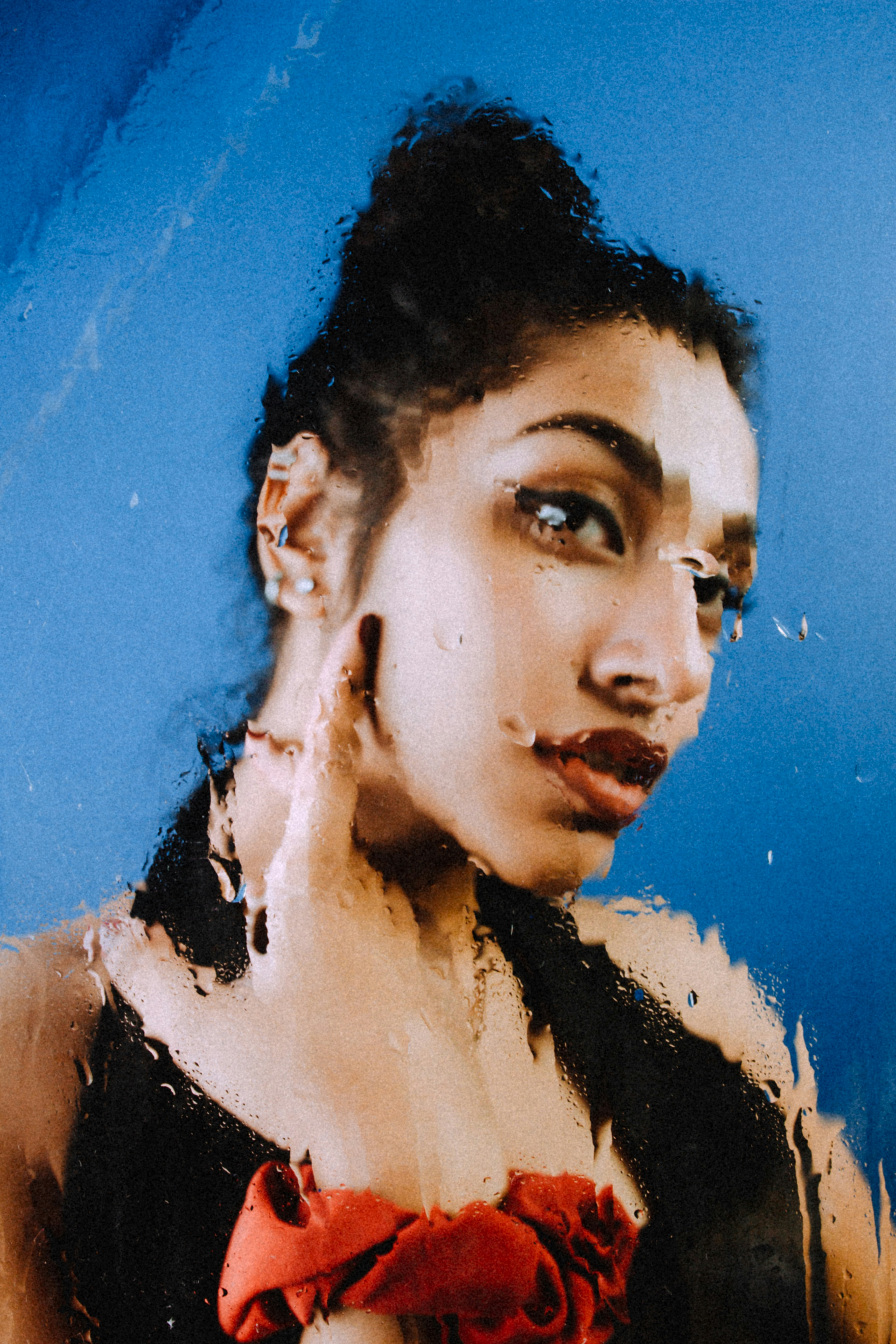 A portrait of a woman partially obscured by condensation on glass, highlighting her expressive gaze against a vibrant blue background.
