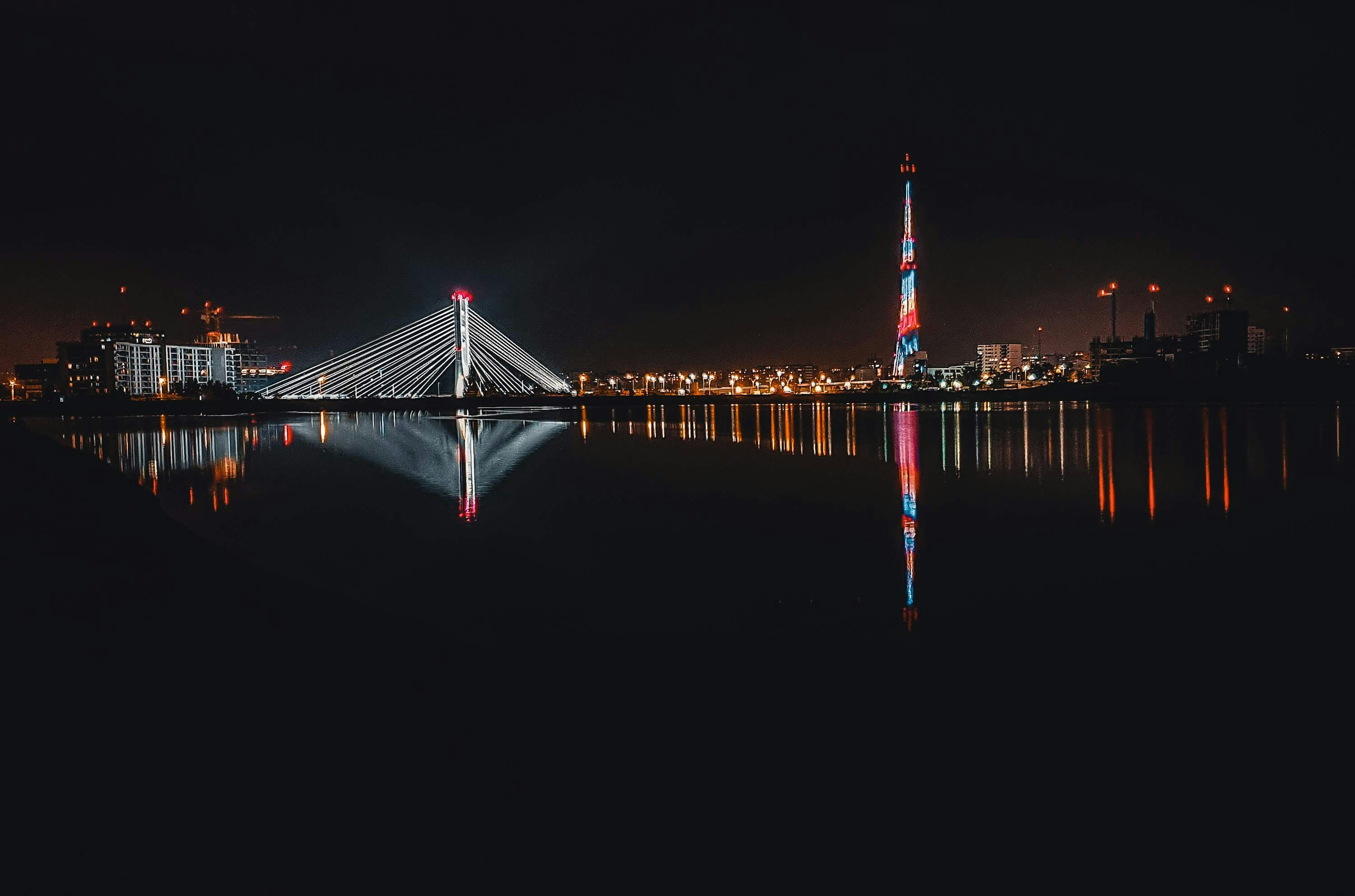 Illuminated bridge and tower reflecting on still water at night, showcasing urban architecture and city lights.