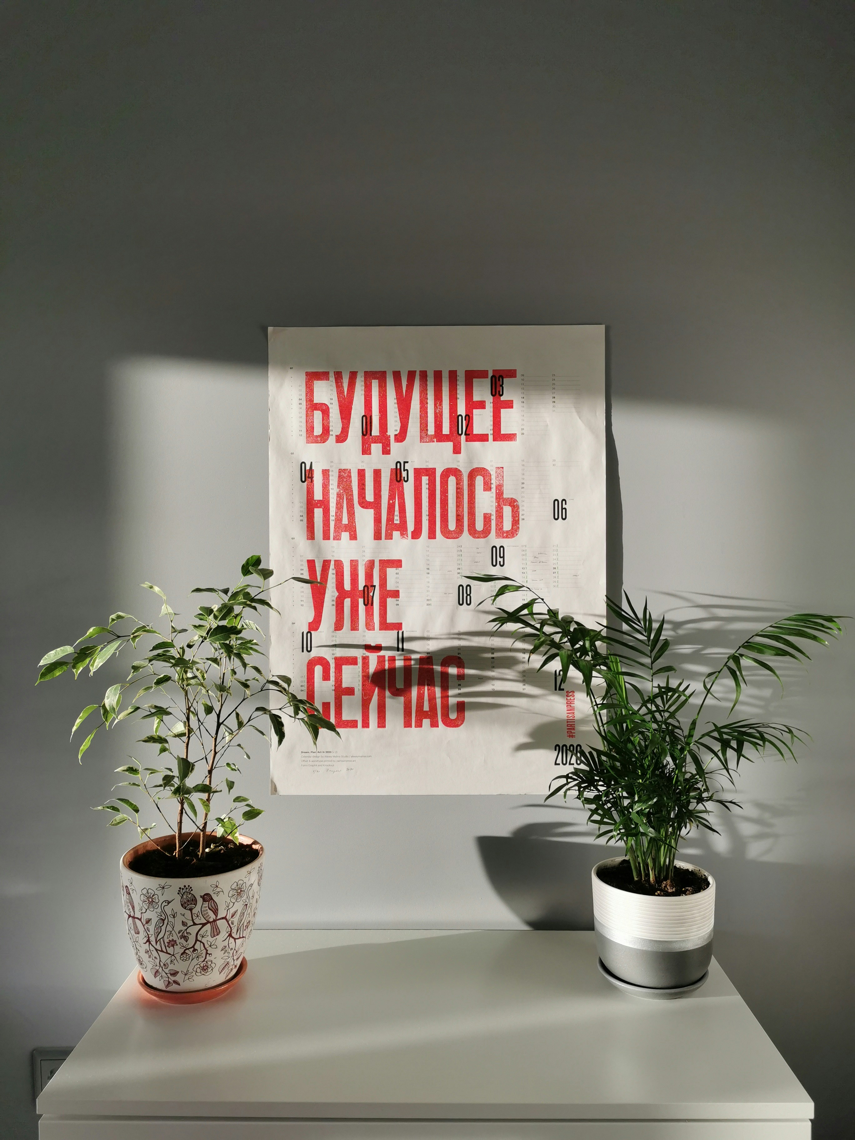 Bold red Cyrillic poster anchors a minimalist still-life scene with two potted plants on a white table.