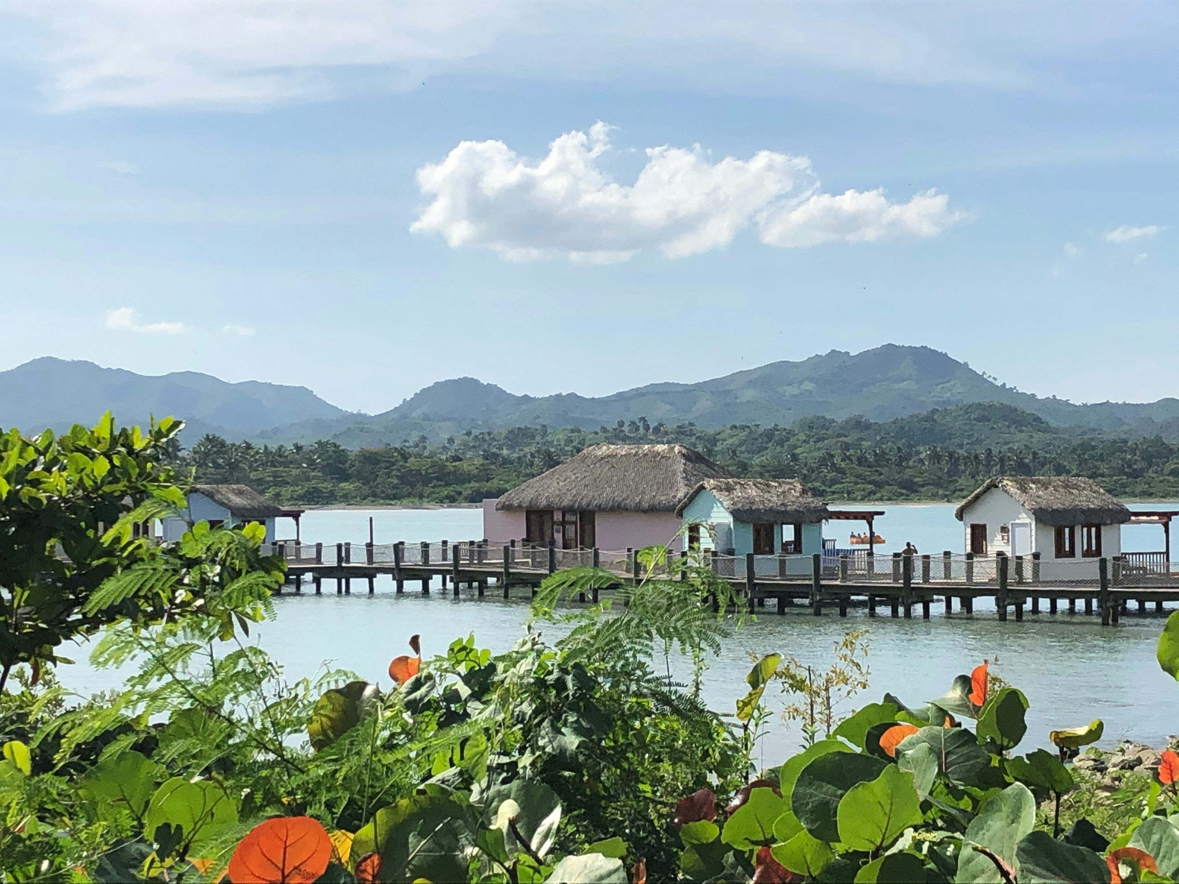 Thatched-roof huts on stilts over a serene lake, framed by lush greenery and distant mountains under a bright blue sky.