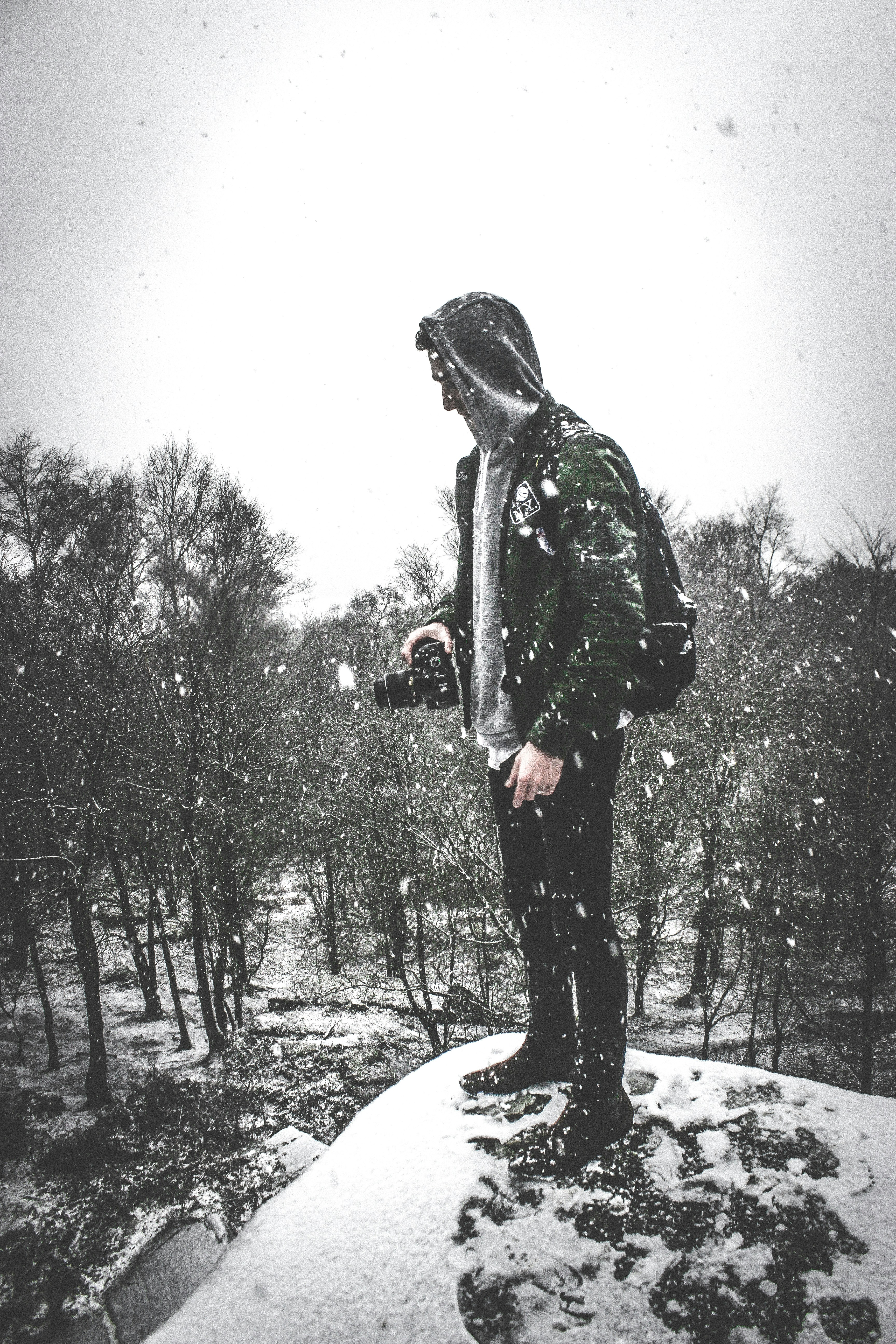 Person standing on a snowy cliff edge during a snowfall, holding a camera.