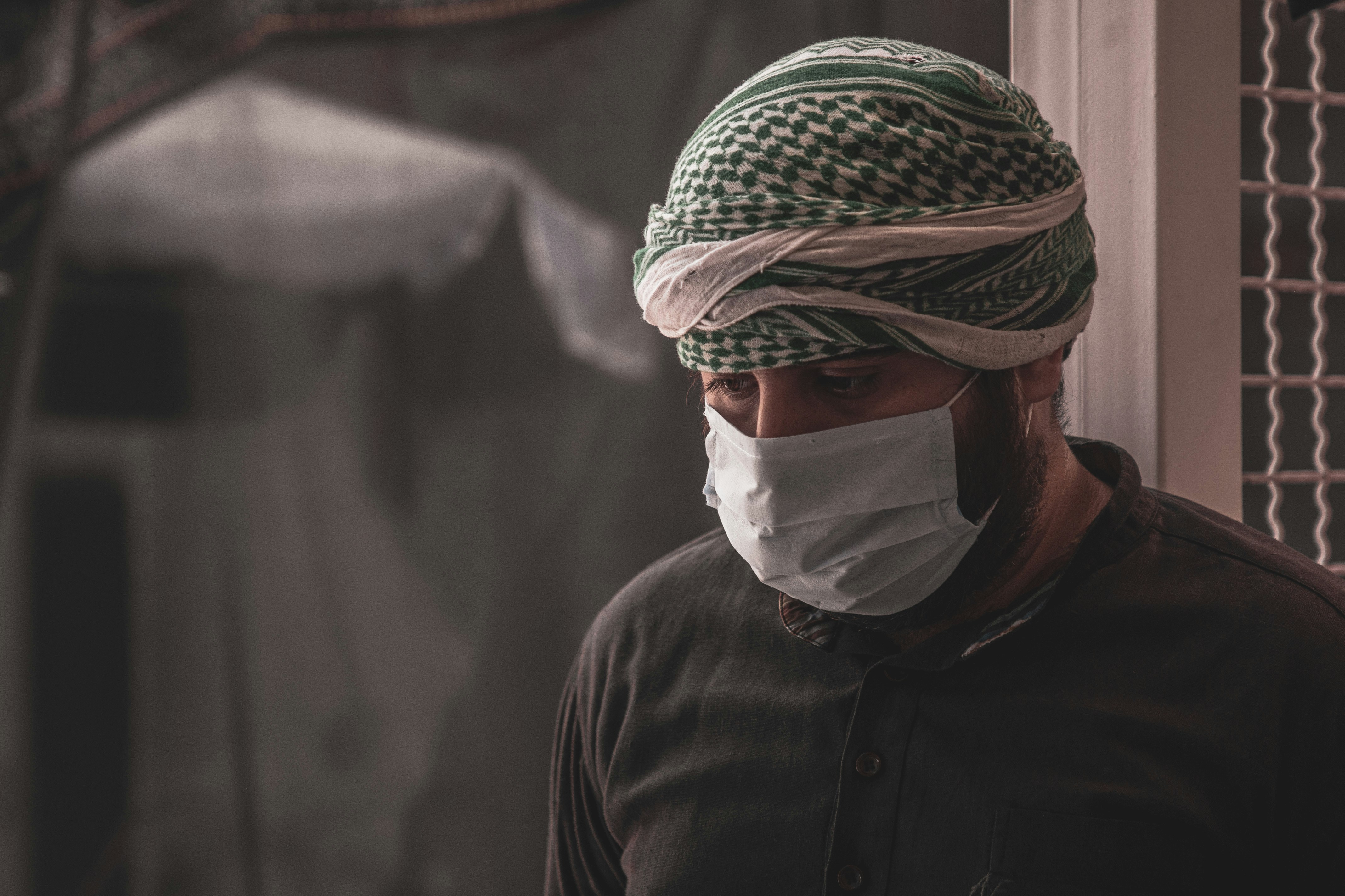 A man wearing a traditional headscarf and a mask gazes thoughtfully, embodying a moment of quiet introspection amidst a blurred background.