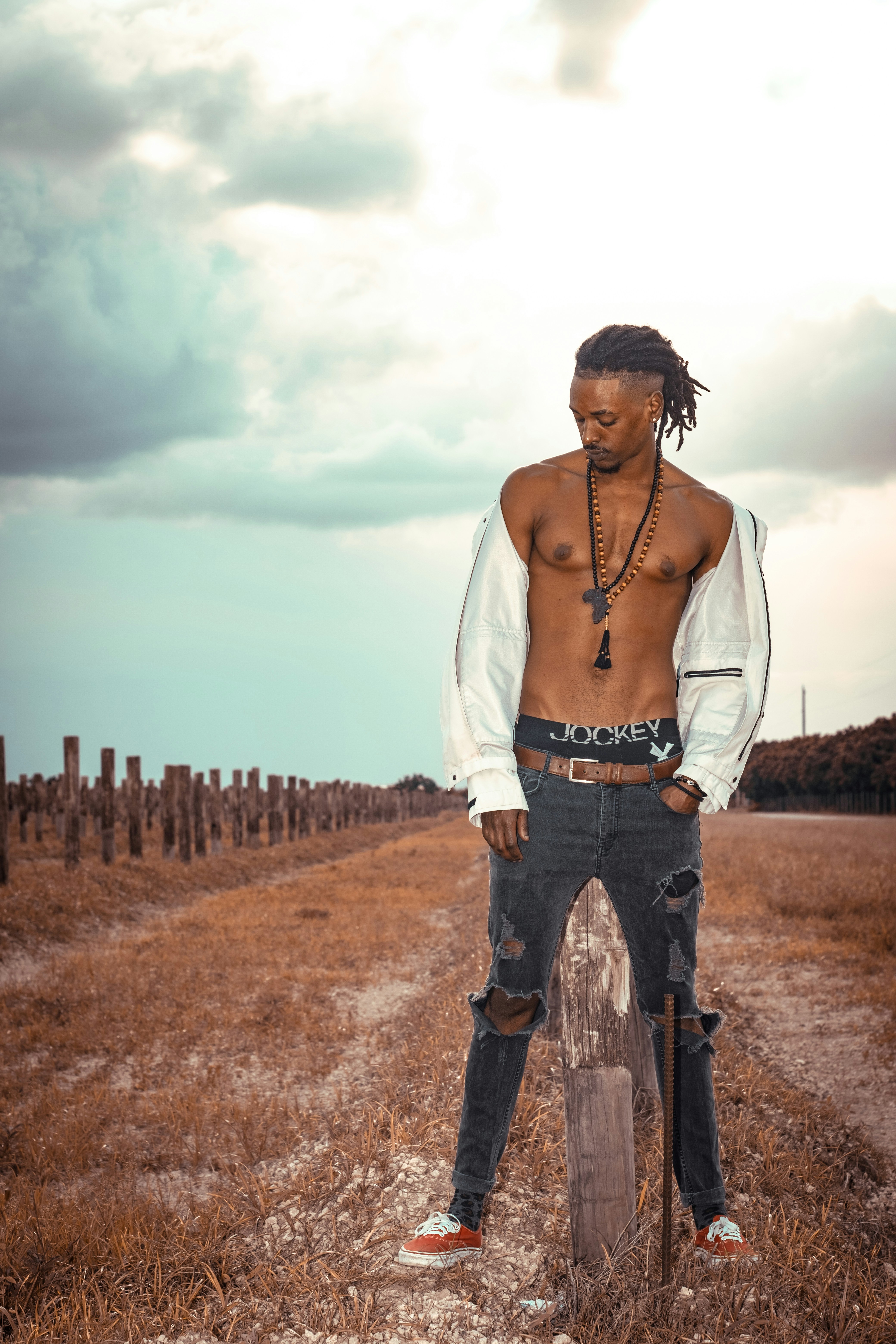 A man stands confidently in a field, showcasing a blend of urban fashion and natural surroundings, with an expressive posture and thoughtful gaze.