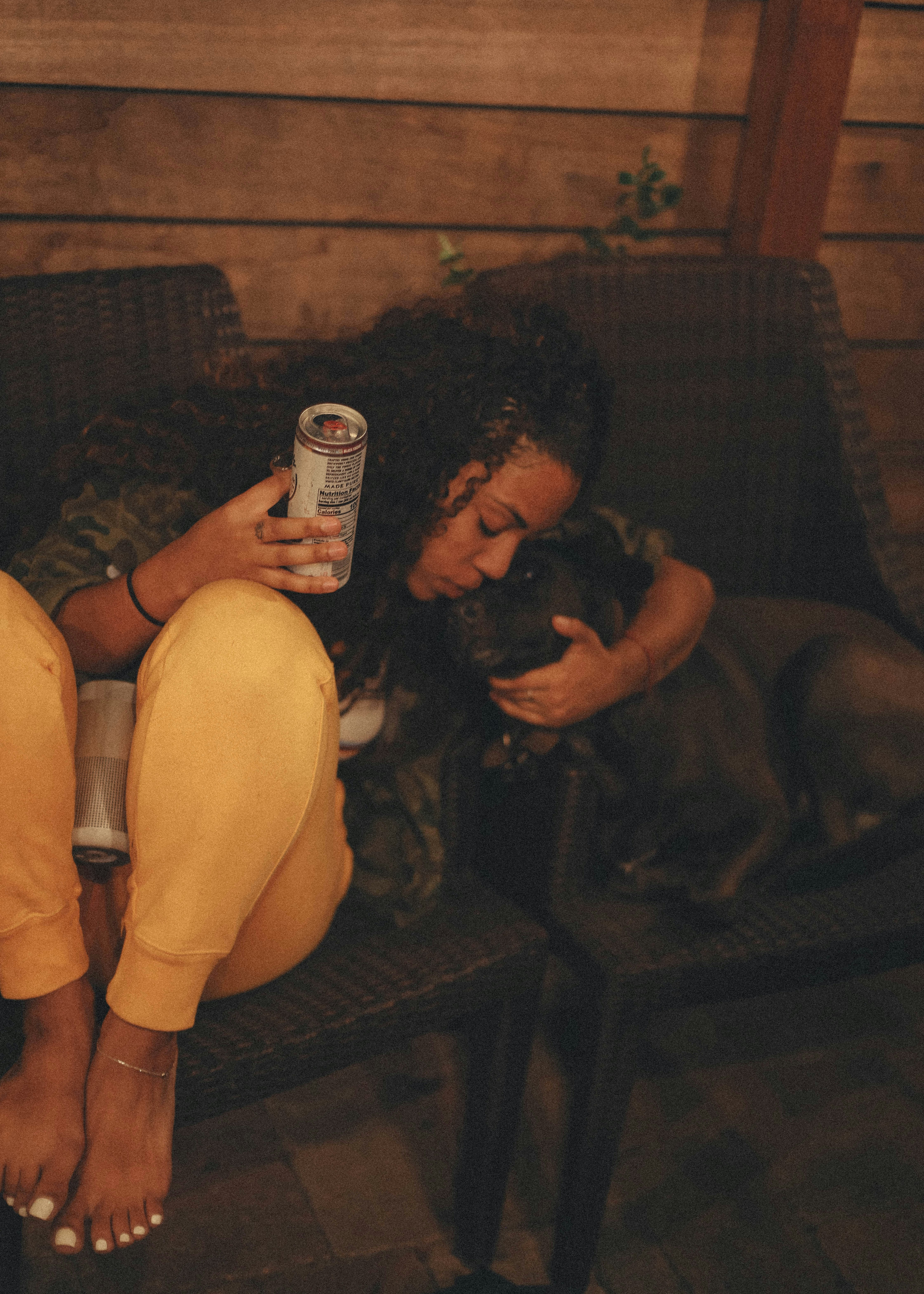 A woman in casual attire shares a tender moment with her dog while holding a drink, seated comfortably in a cozy outdoor setting.