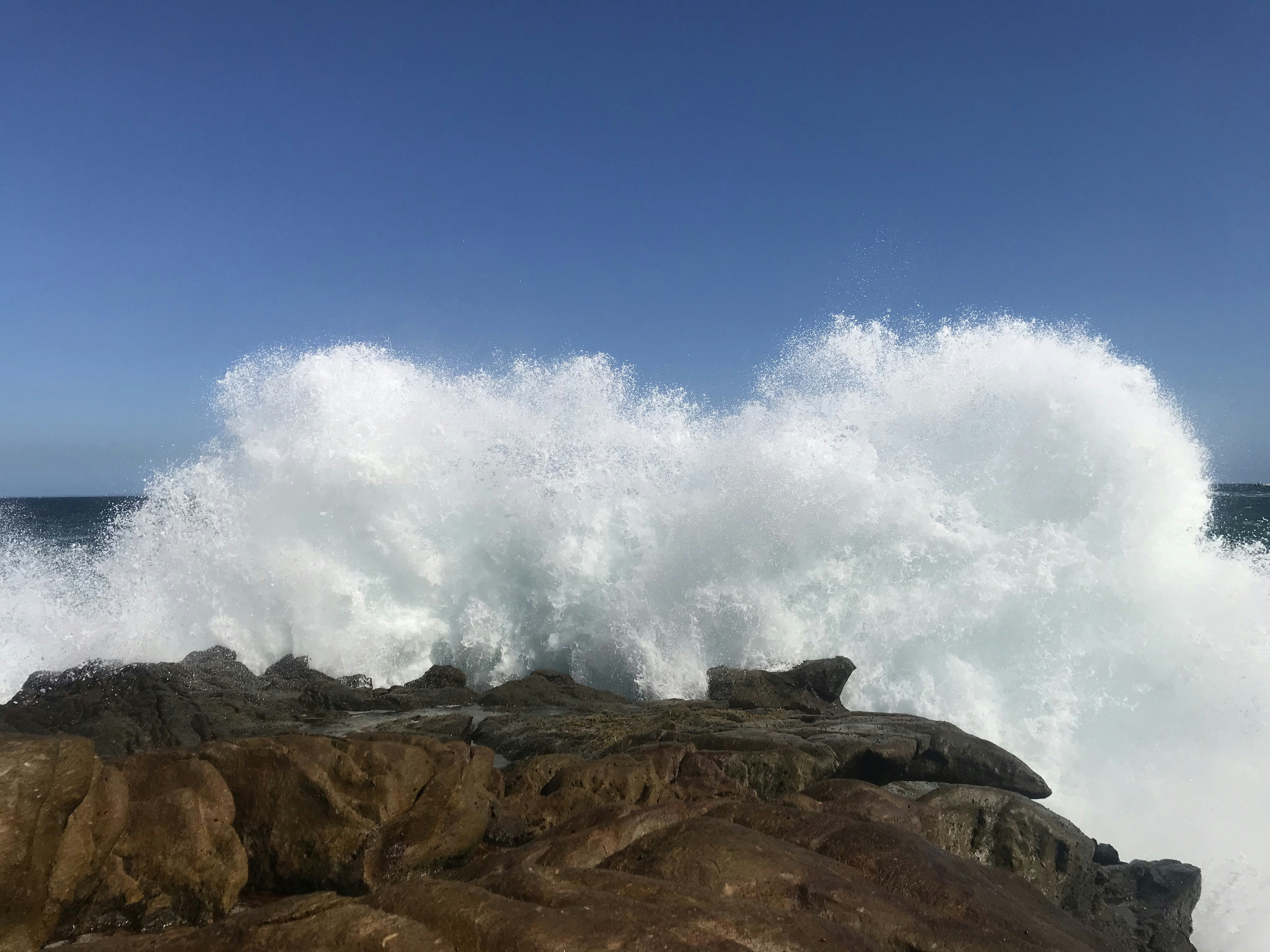 A powerful wave crashing against rugged rocks under a clear blue sky.